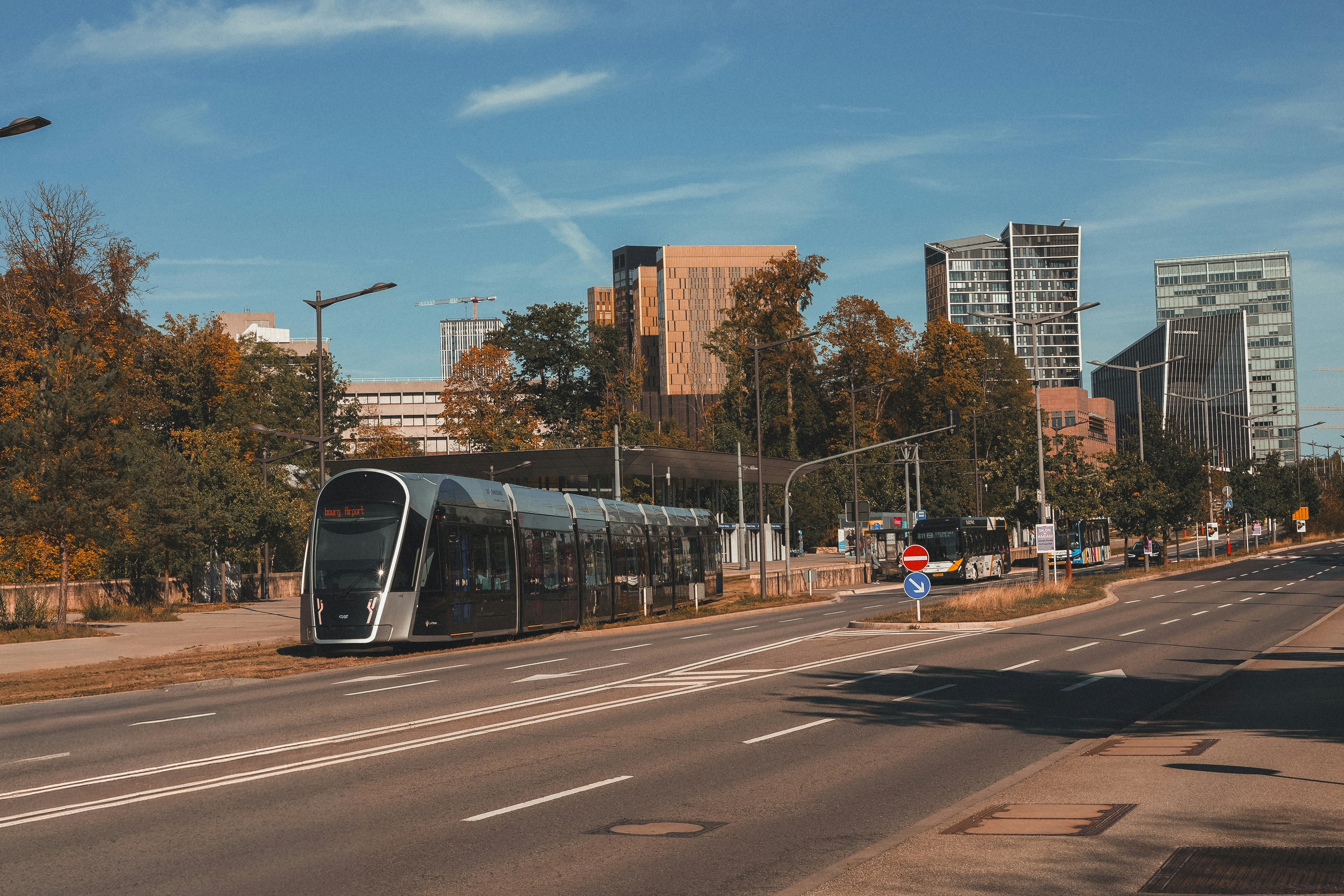 Autumn city street with a sleek tram passing along the tracks; modern office towers rise in the background under a bright blue sky.