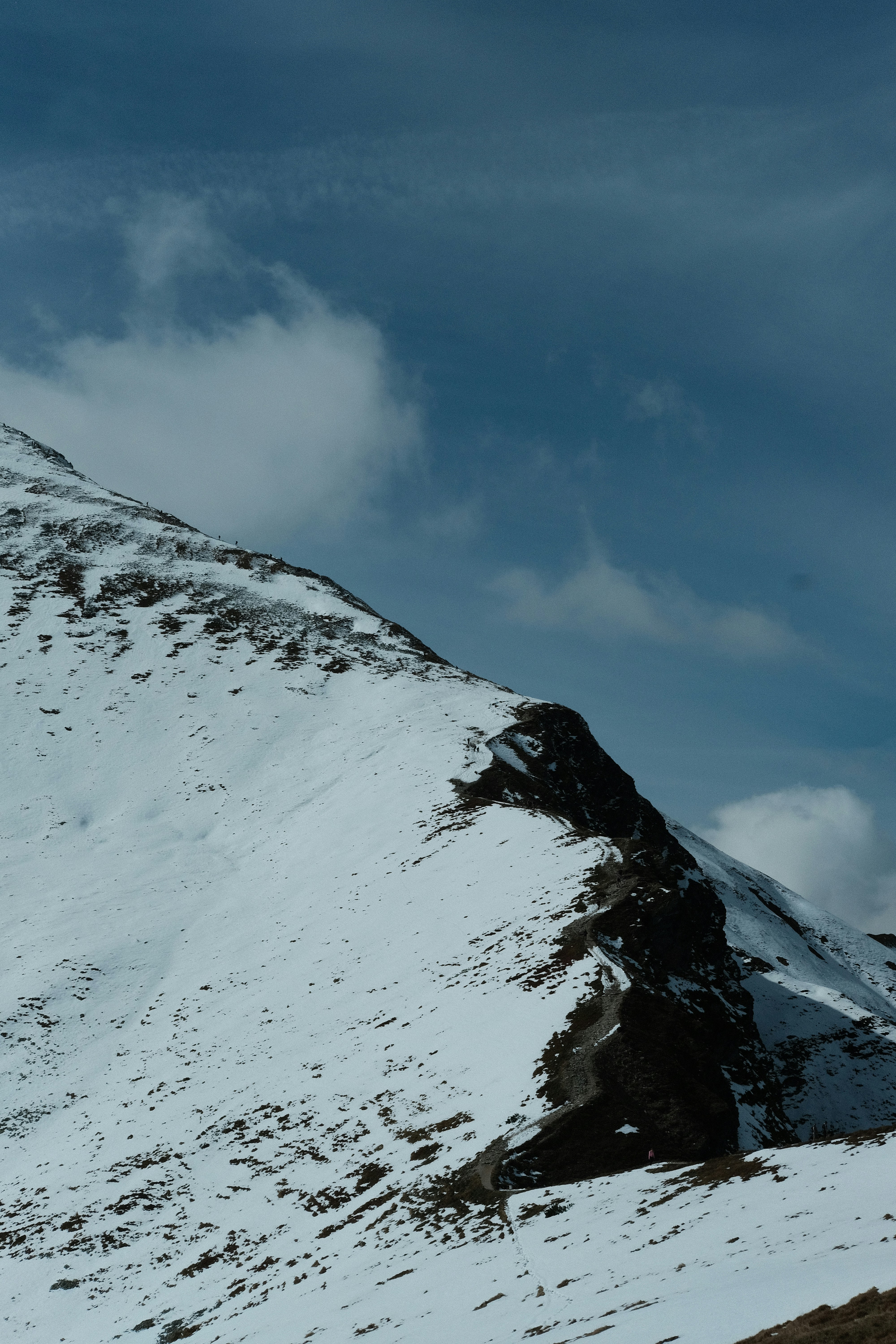 Snow-covered mountain ridge against a blue sky, with a dark rocky spine and scattered clouds. A landscape shot emphasizing the contrast between snow and rock.