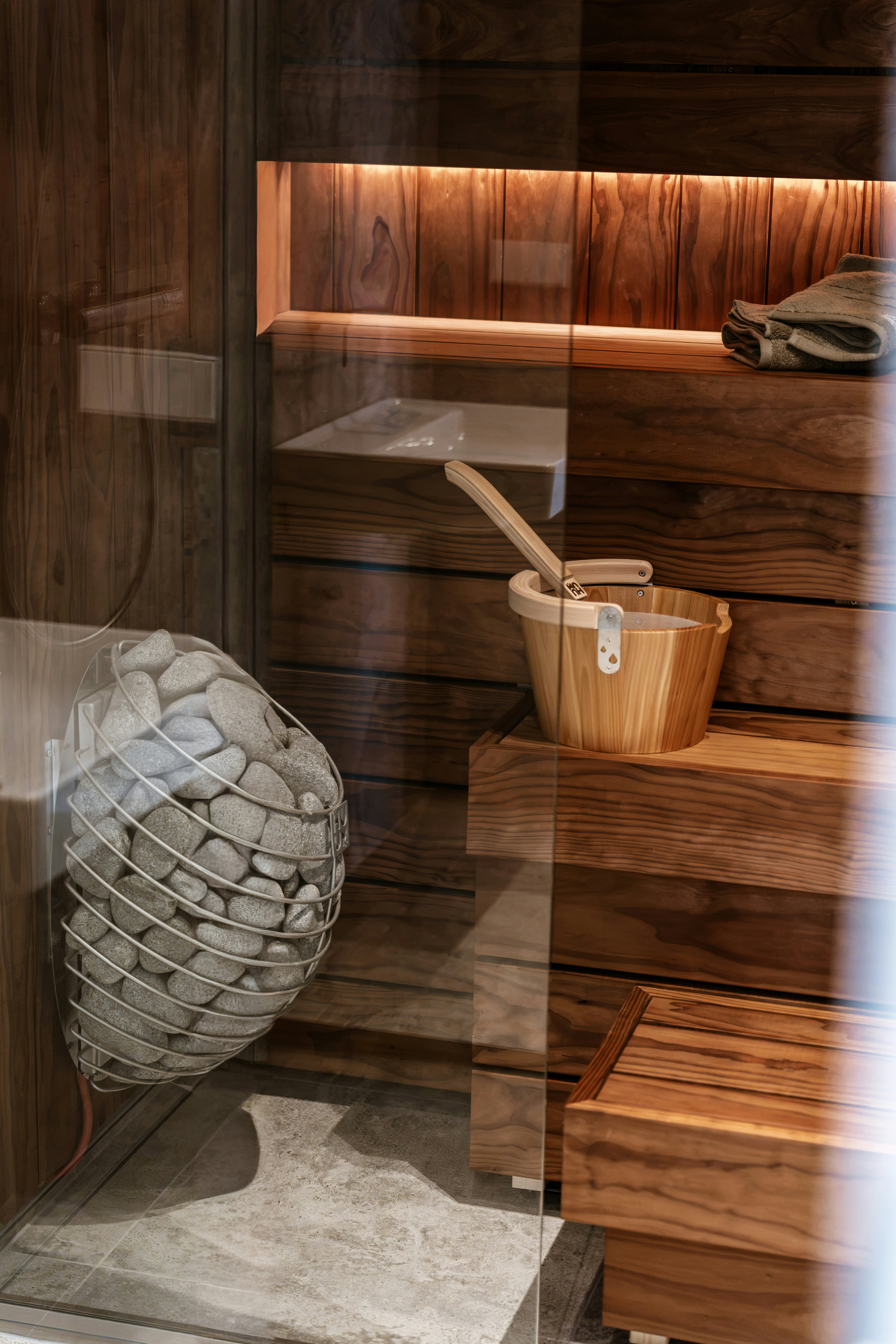 Wood-paneled sauna interior with a lit niche, a wooden bucket and ladle, and a textured stone floor; soft lighting and a towel on the bench emphasize a serene spa setting.