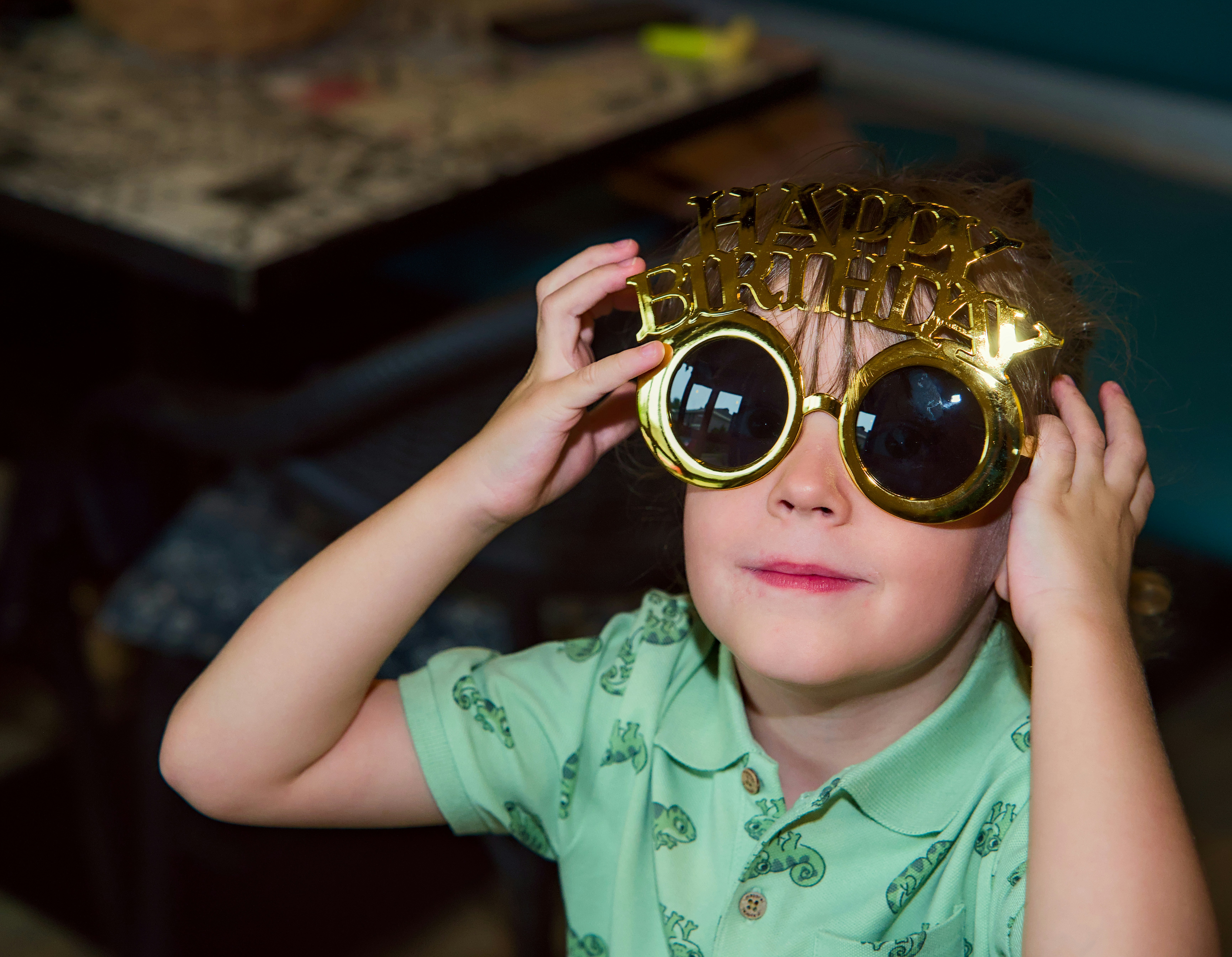 Playful child wears oversized gold party glasses, a festive mask reflecting light, in a warmly lit indoor setting.