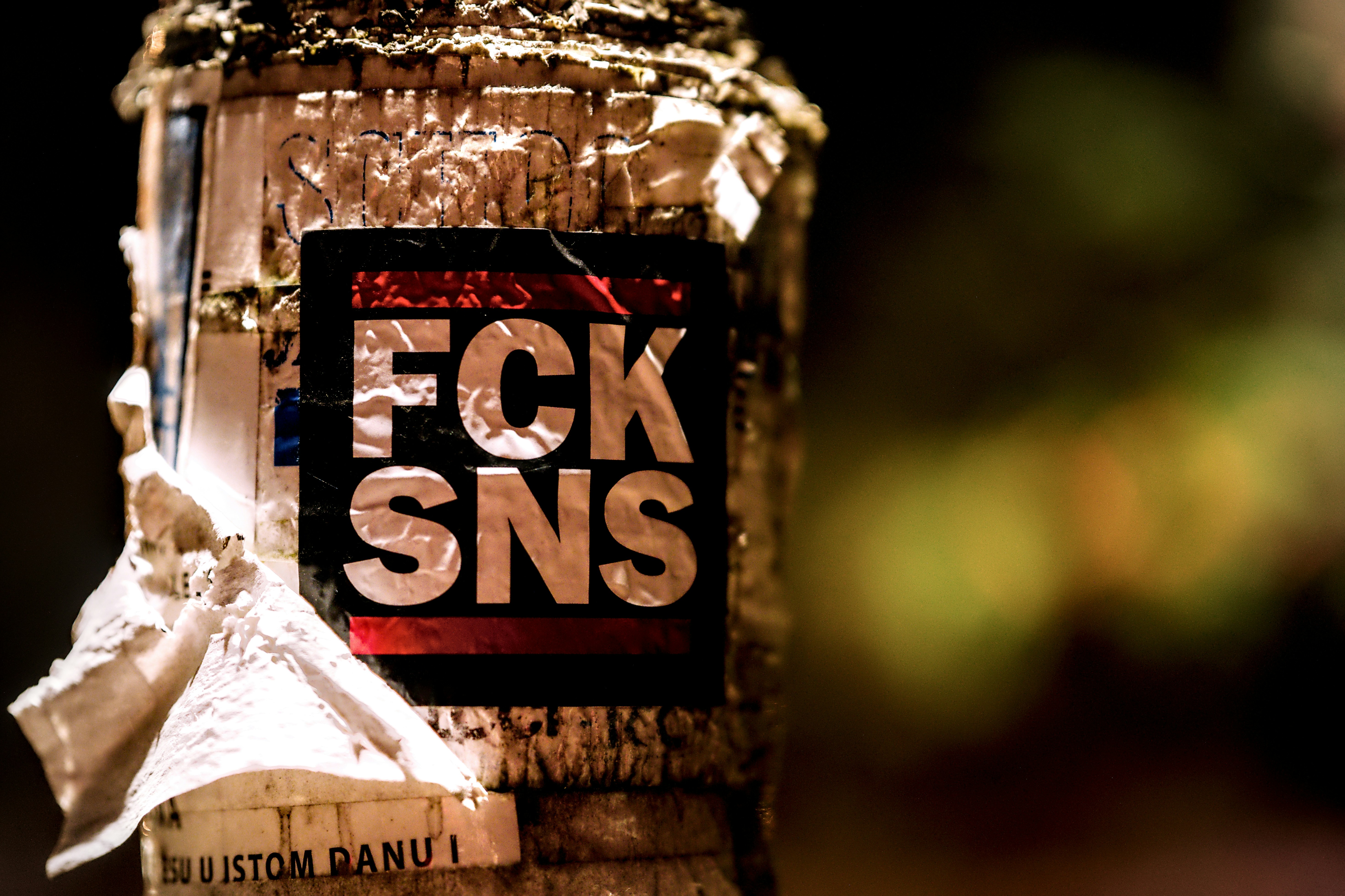 Tag sticker on a weathered trash can with peeling tape; the label reads FCK SNS and stands against a blurred, warm-toned background.