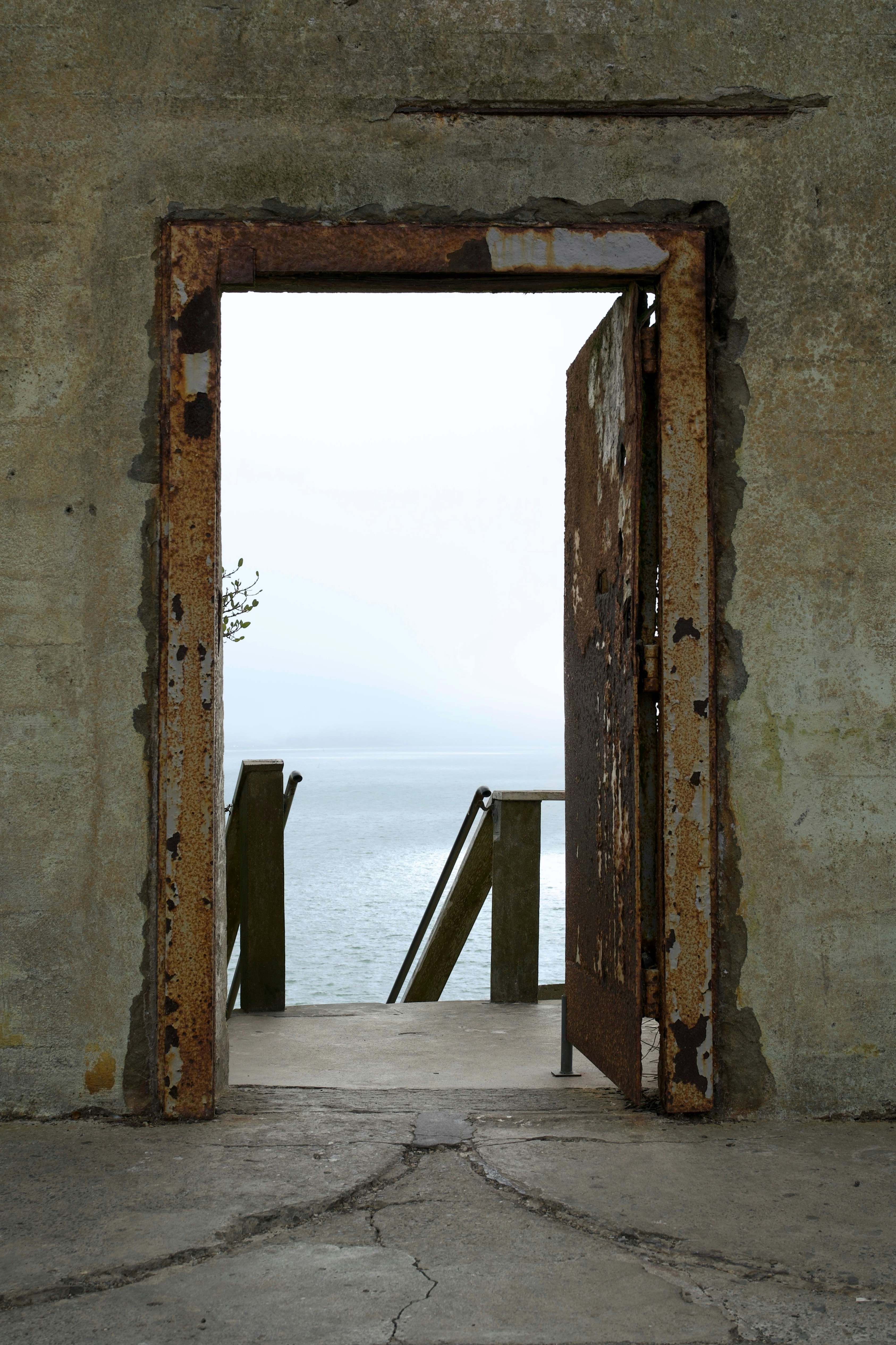 Rust-streaked doorway opens to a calm sea beyond a concrete threshold; weathered metal railing and steps hint at a ruined vantage point above the water. A minimalist view through an industrial frame reveals distant horizon and soft light.