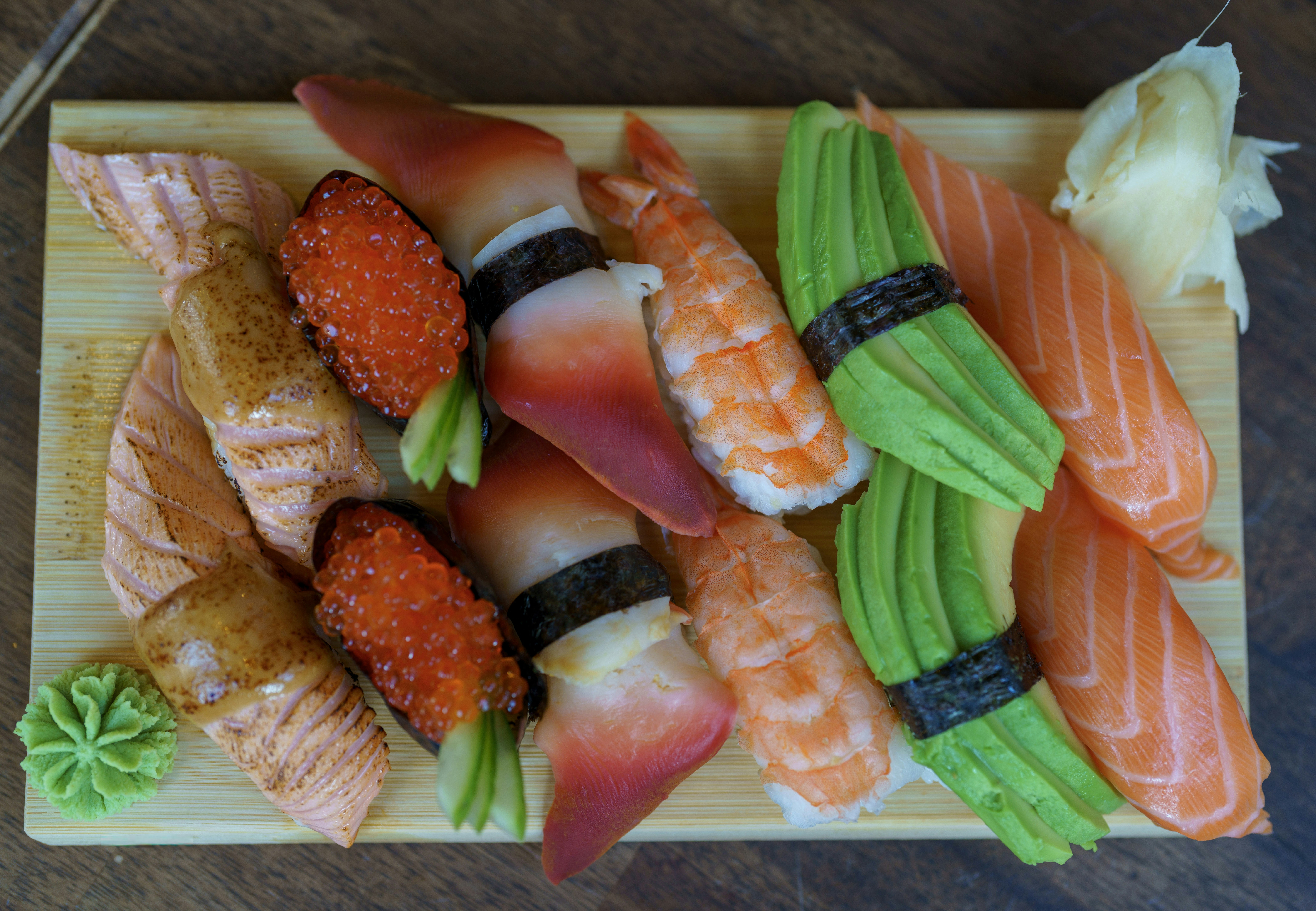 Colorful sushi assortment on a wooden board with salmon, tuna, shrimp, and avocado-wushi alongside wasabi and pickled ginger.