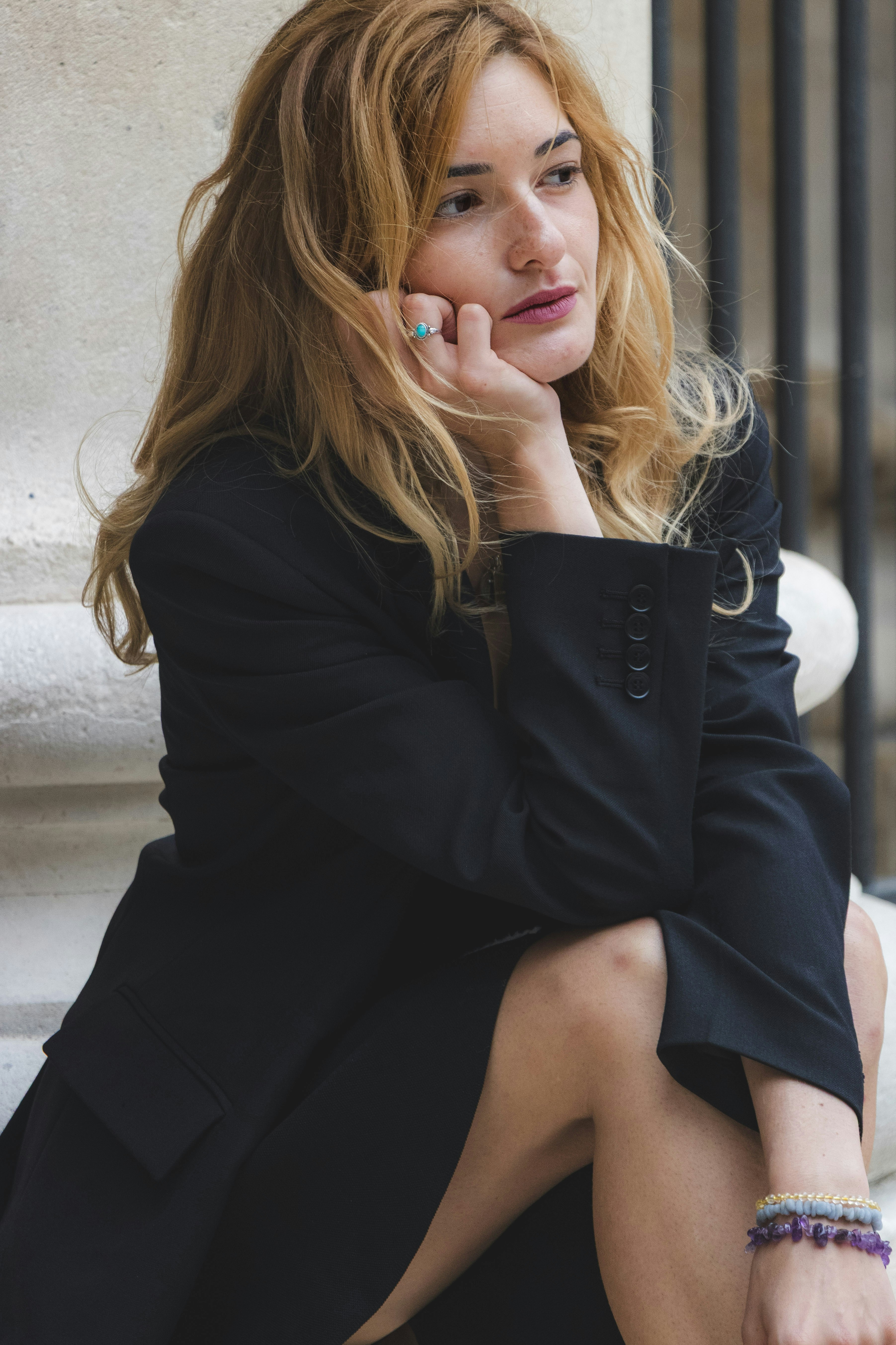 A woman sits against stone architecture, resting her chin on her hand; black blazer, blonde hair, and colorful bracelets add contrast to the muted backdrop. This is a candid fashion portrait.