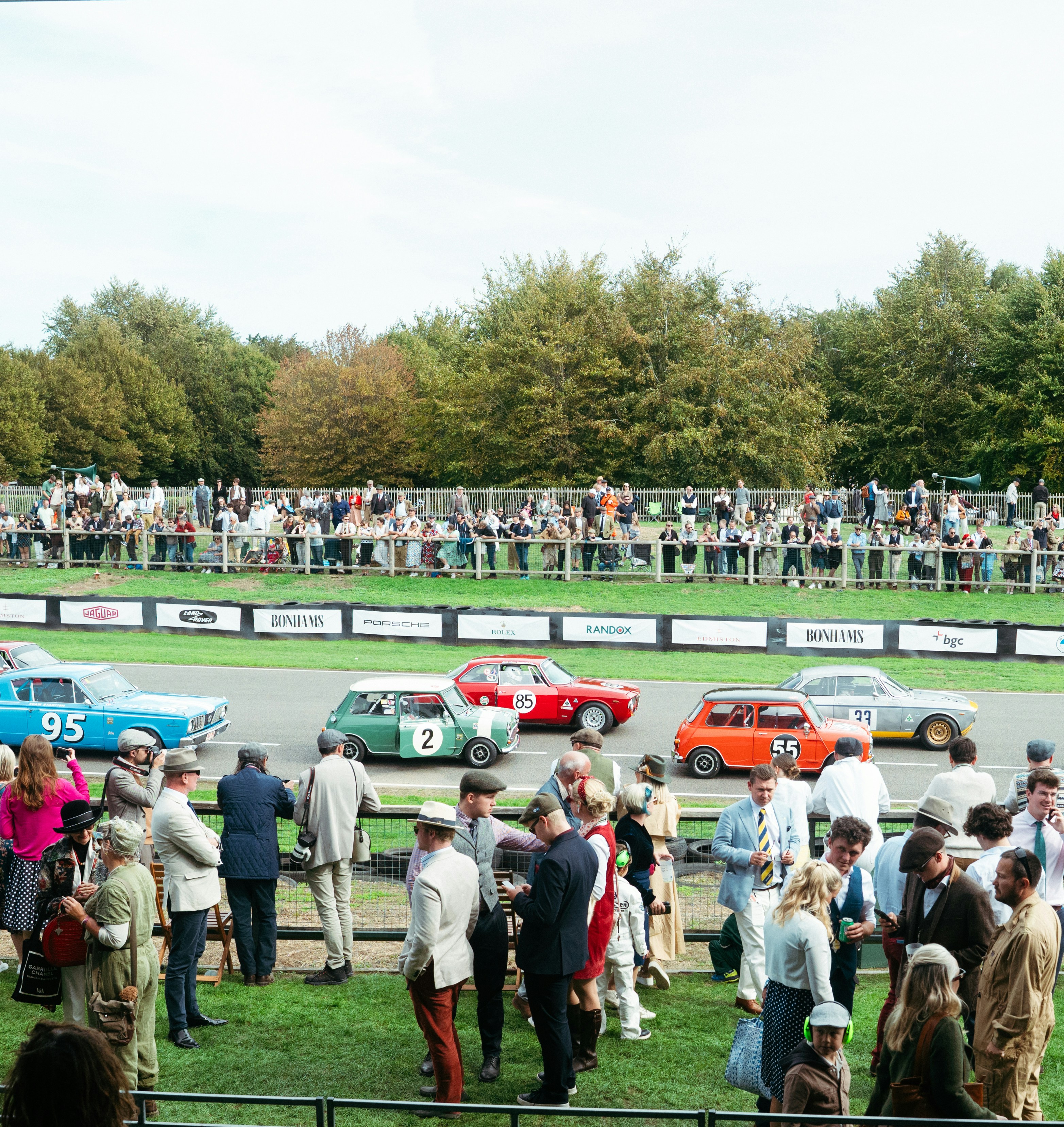 A vintage car race day scene with a row of miniature race cars on a track and a crowd of spectators in the foreground. The image captures a lively event with tight composition and classic automotive color accents.