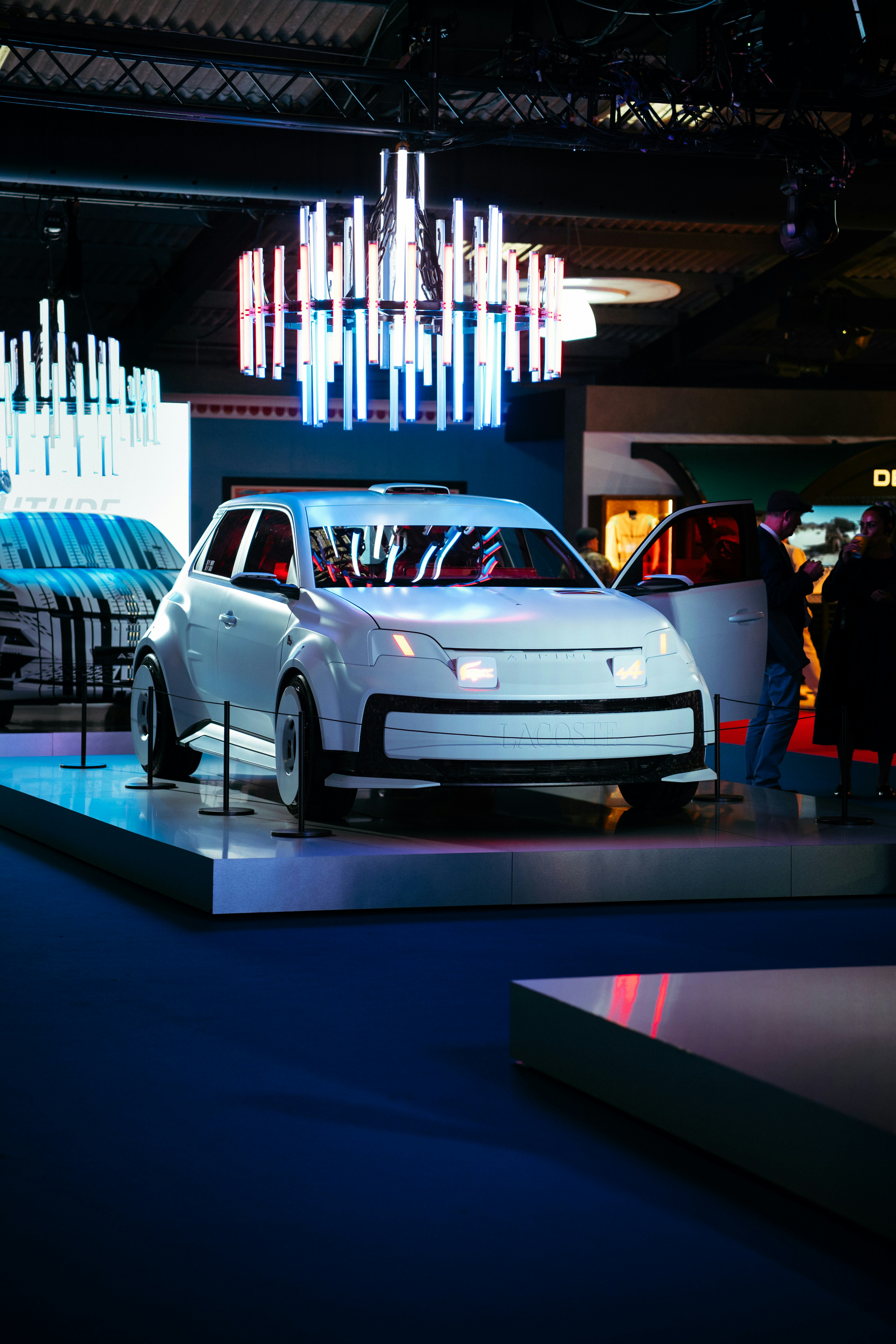 A white concept SUV is spotlighted on a raised platform at an auto show, surrounded by neon-lit decor and visitors in the background.