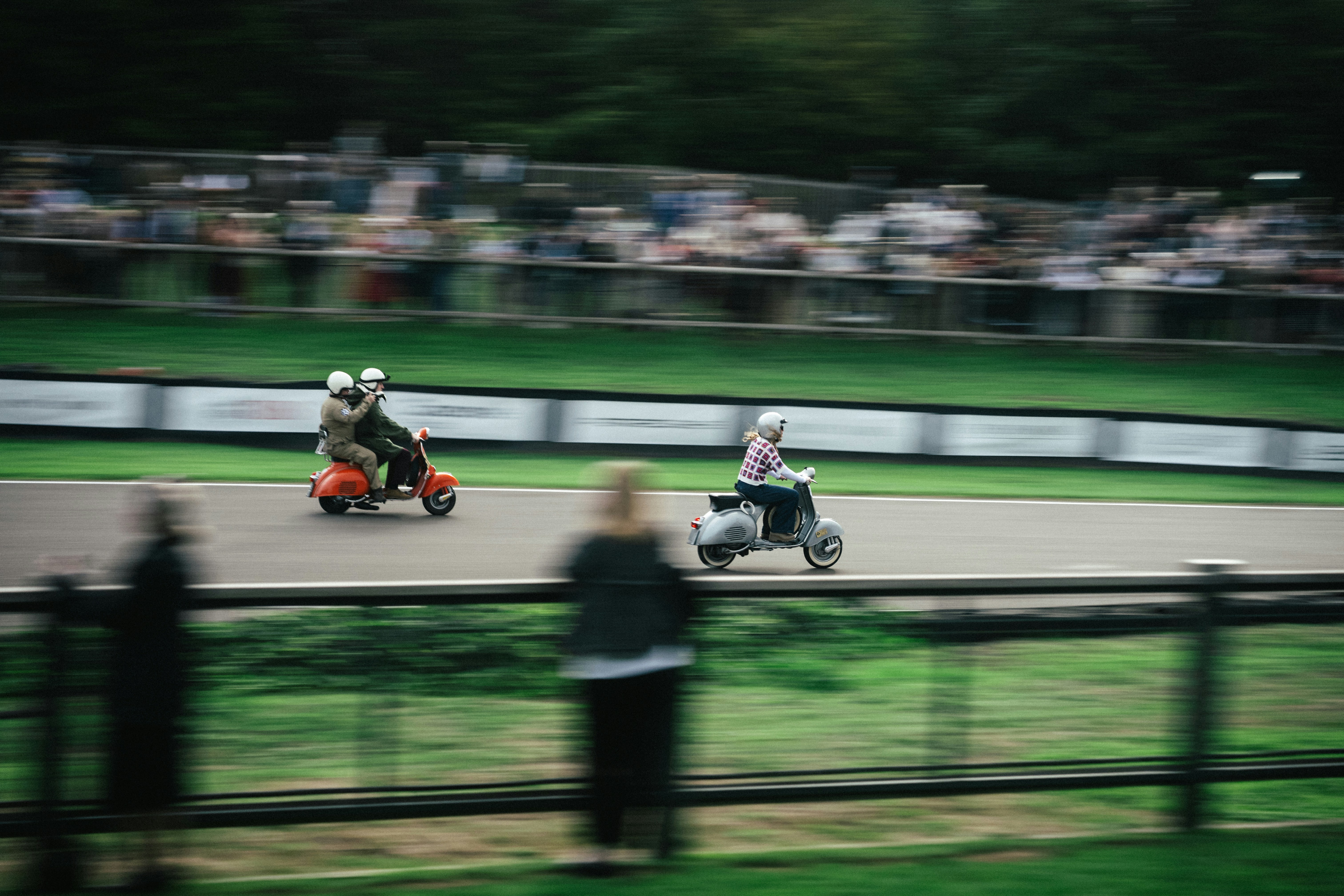 Two vintage scooters race along a track with bystanders and blurred foreground, emphasizing motion and competition.