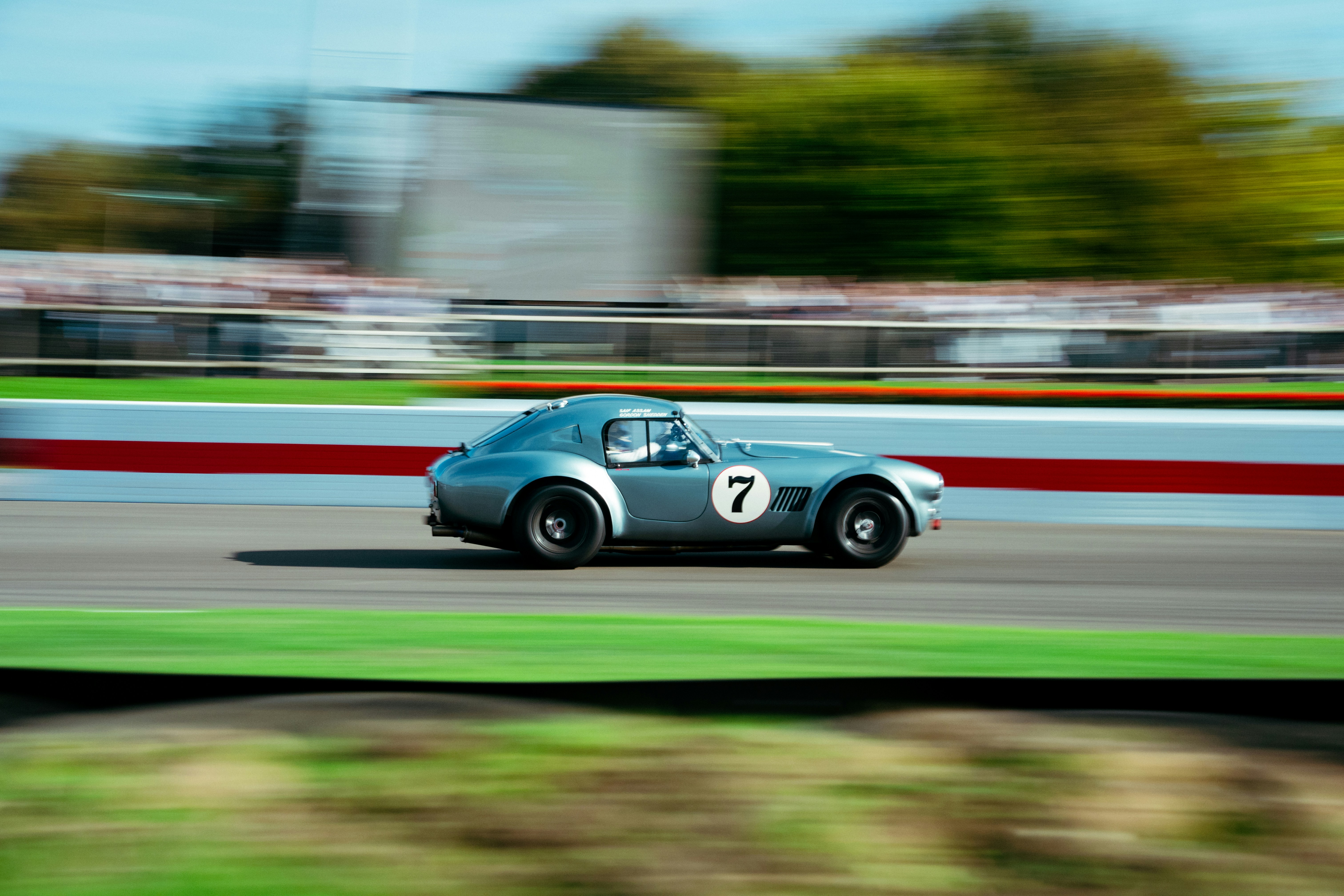 A classic blue race car with number 7 speeds along a track, captured with motion blur to convey acceleration and motion; the scene emphasizes the vehicle and race backdrop.