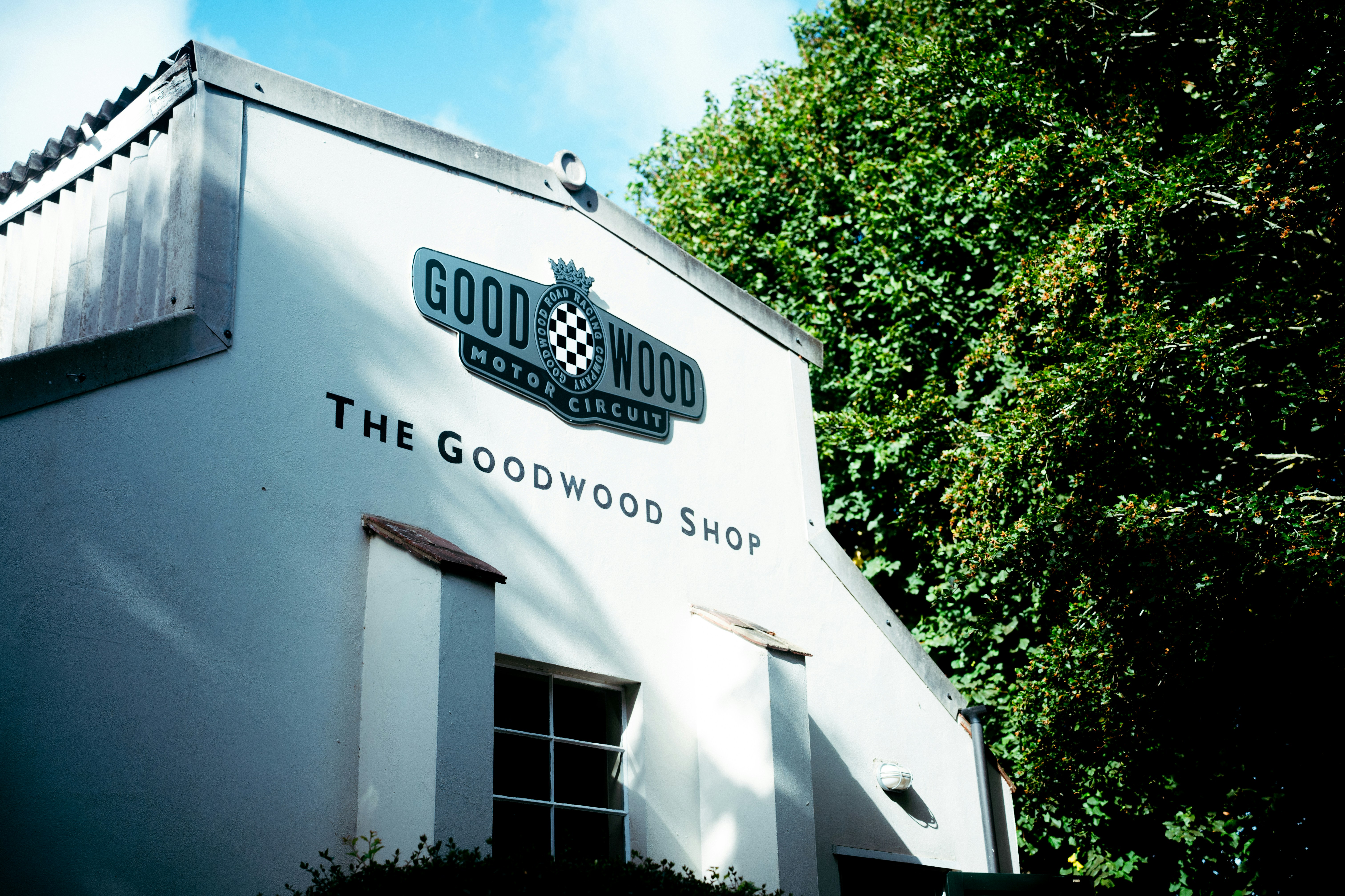 White building with Goodwood Motor Circuit signage, partially shaded by greenery, capturing a vintage shop facade.