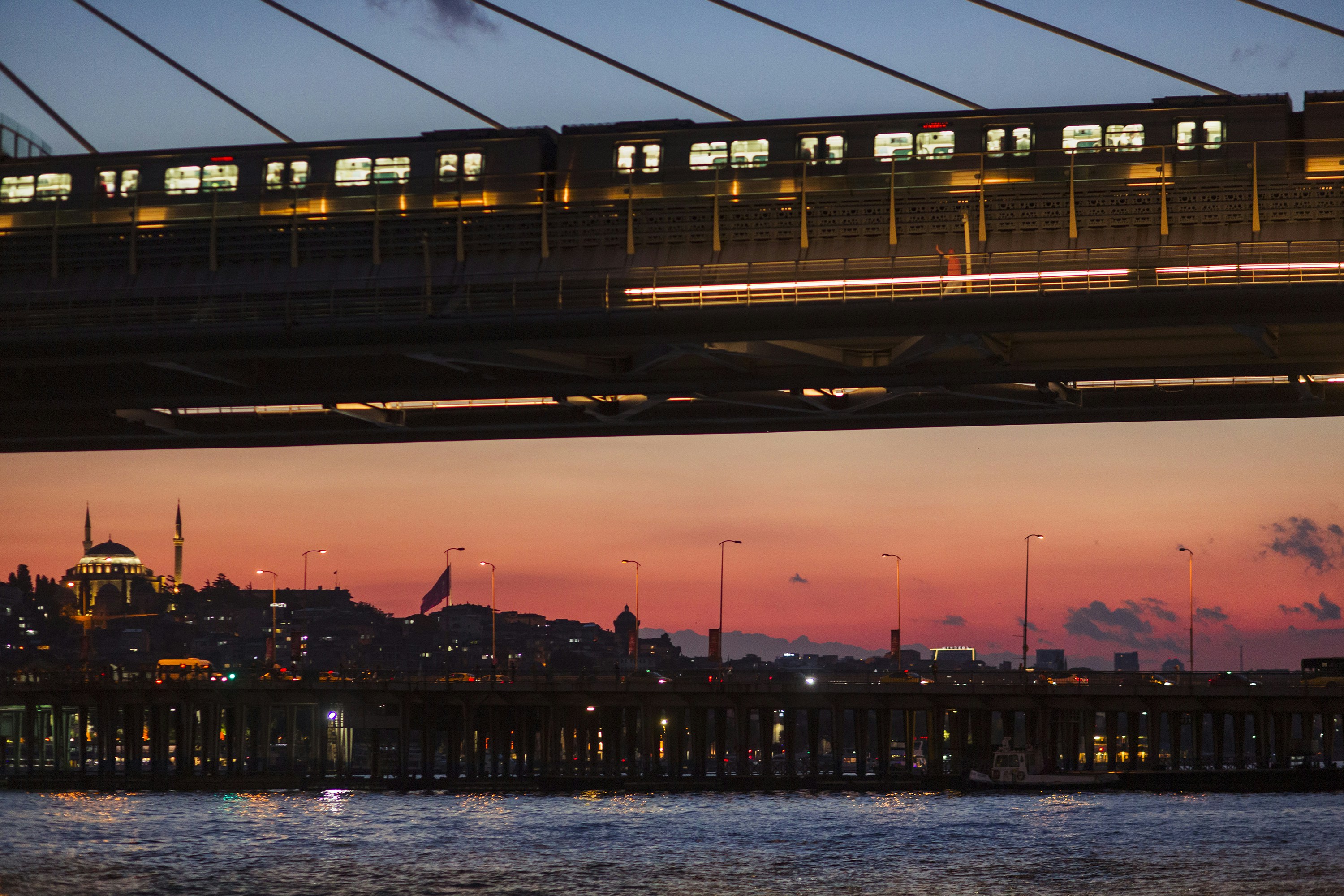 Dusk light silhouettes a long bridge with a train passing overhead over a calm harbor, city skyline fading to twilight in the background.
