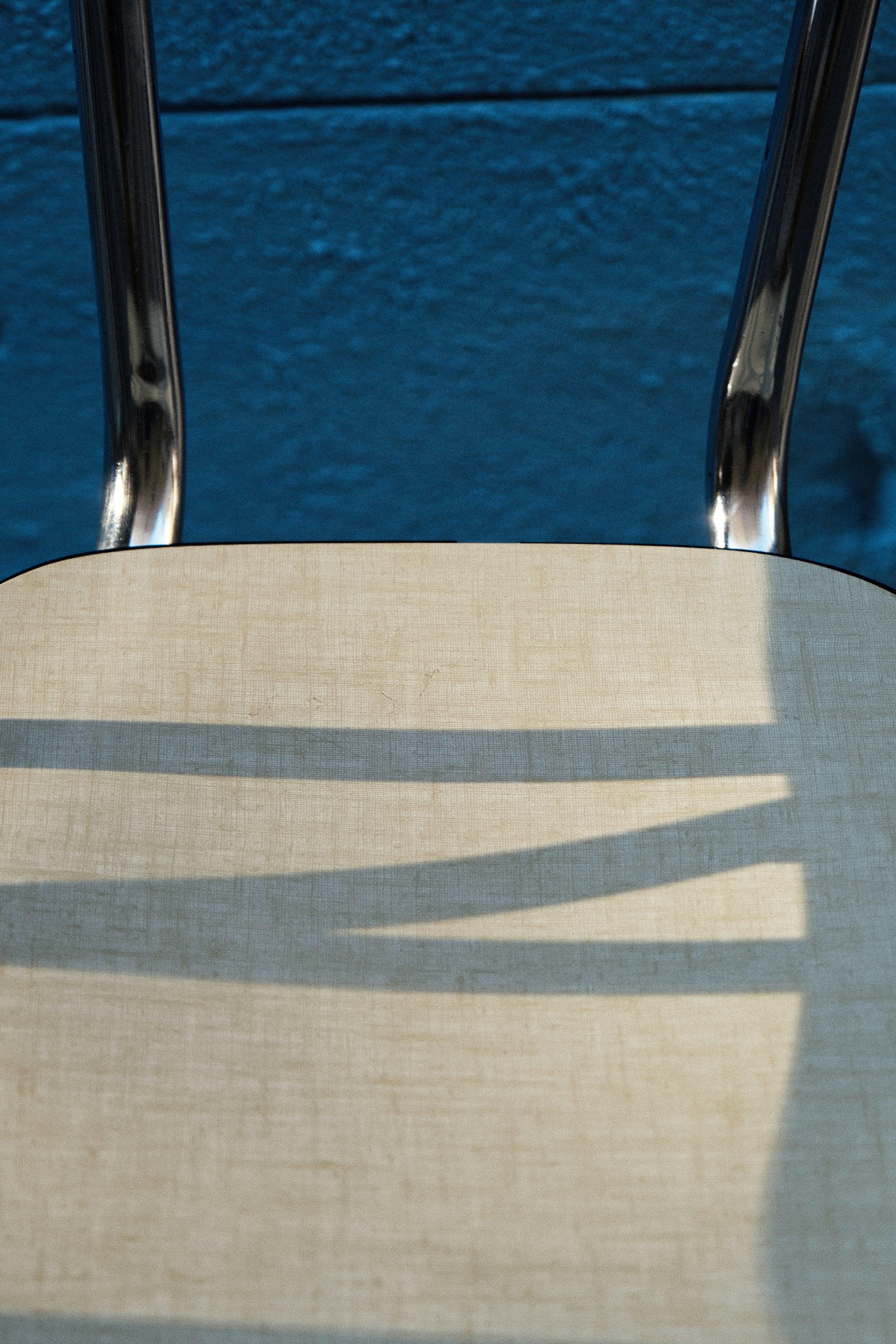 A sunlit beige fabric table surface with bold chair shadows and chrome legs against a blue wall, capturing geometric light and texture. This is a simple still life scene with metallic accents.