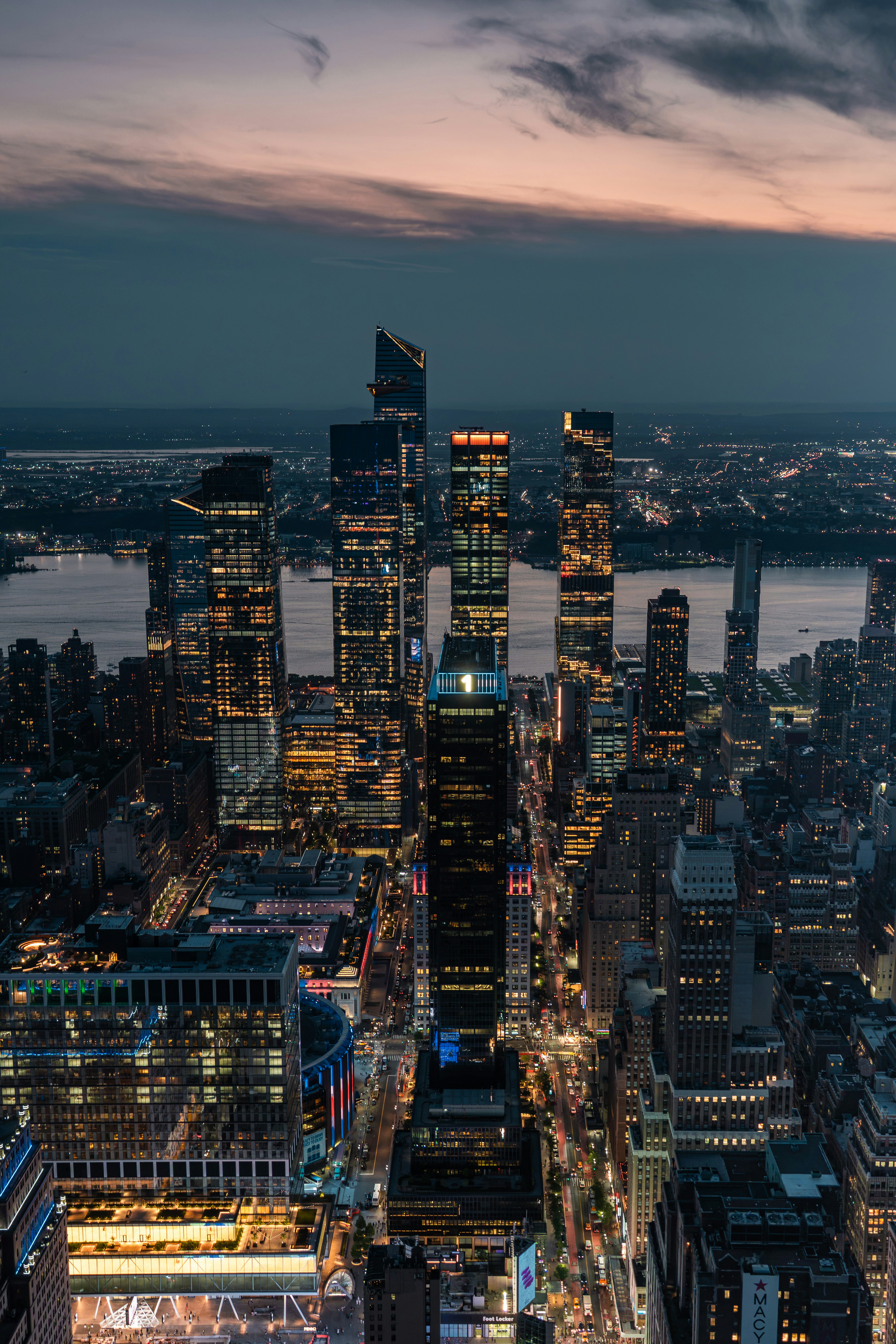 City skyline at dusk with illuminated skyscrapers lining a river, viewed from above as streets glow with traffic. Aerial perspective highlights the vertical geometry of the dense downtown core.