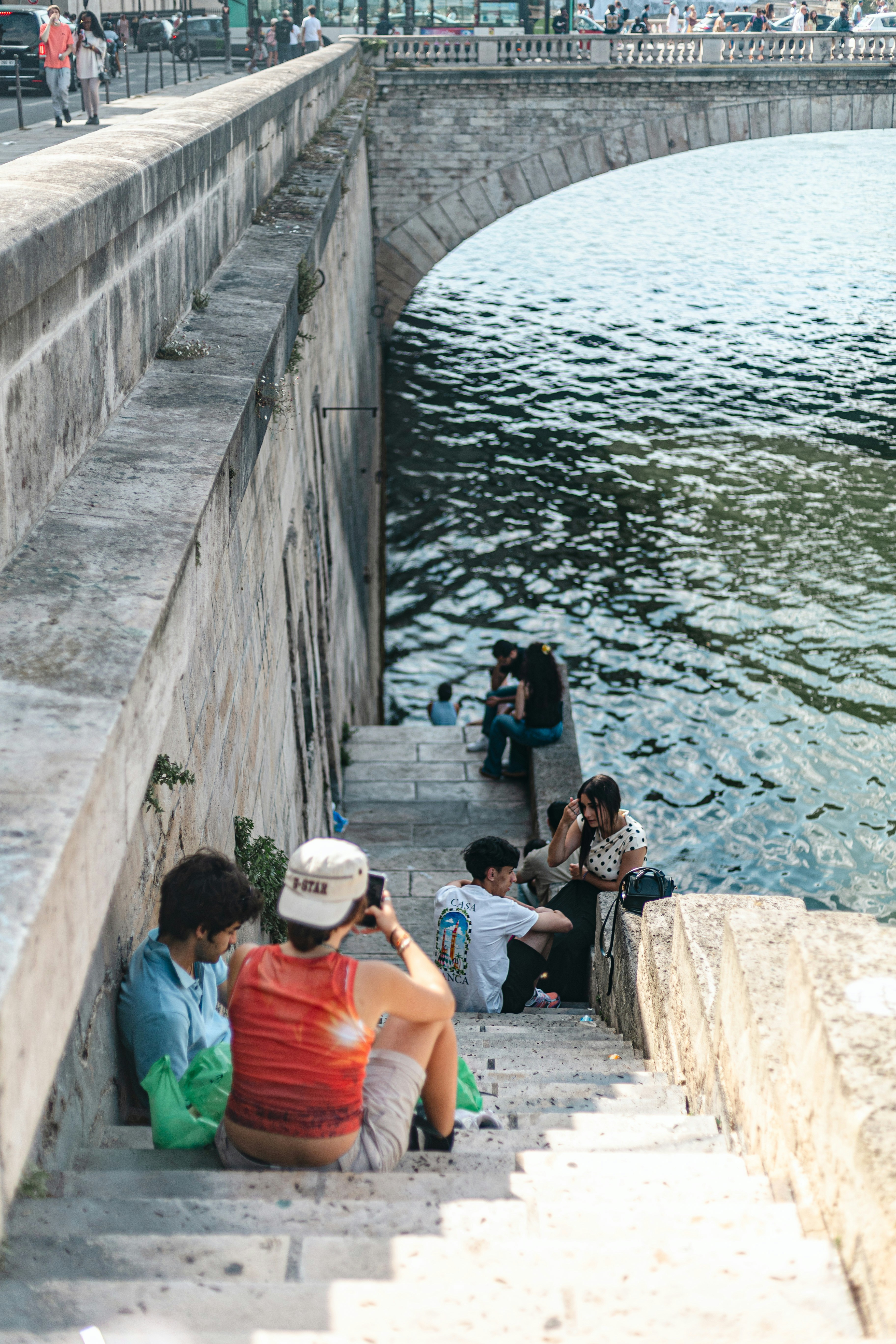 Groups of people sit on stone steps beside a calm river, with a stone wall and arching bridge above them. The scene captures casual urban leisure along a waterfront, including a person in a red top and a man wearing a hat taking a photo.