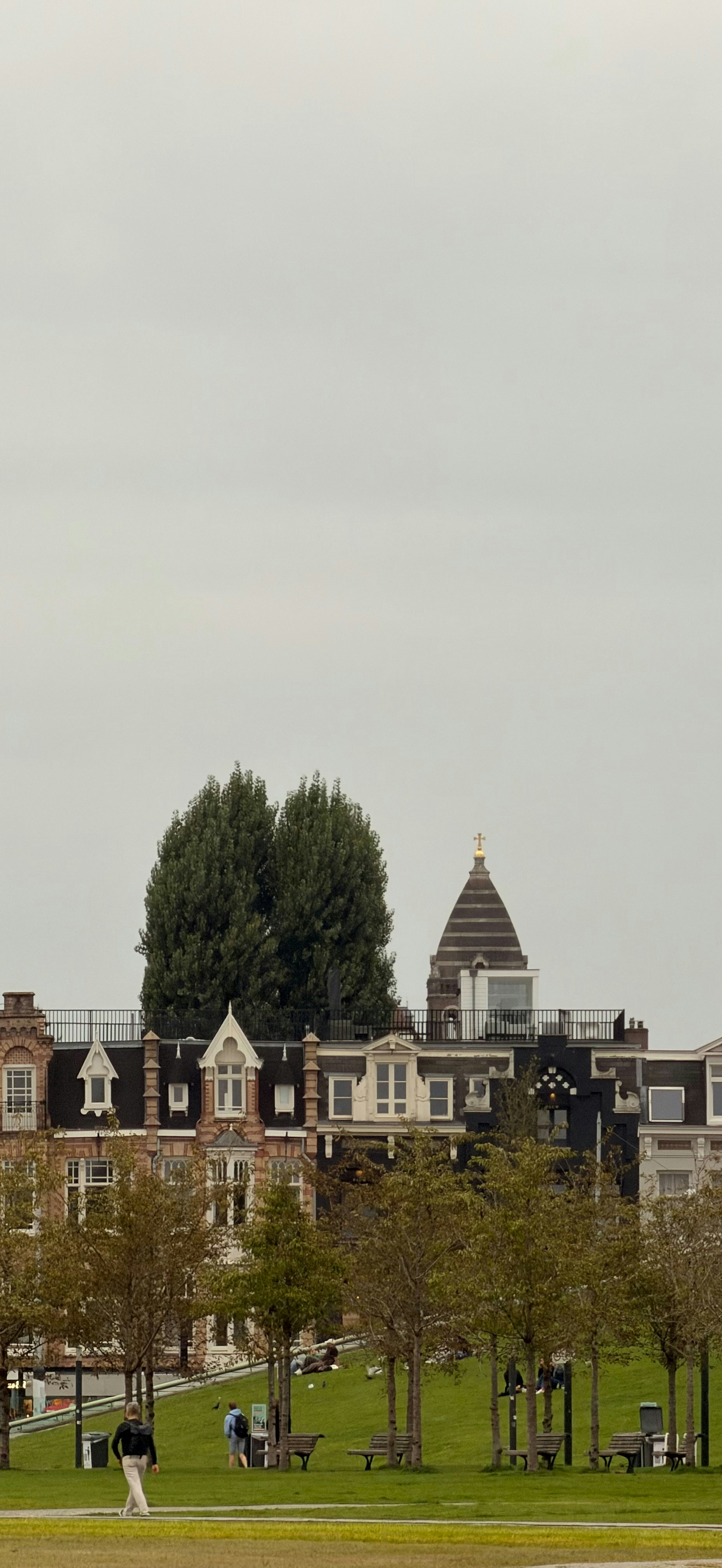 A row of ornate townhouses sits along a grassy park with trees and a few pedestrians, under a pale overcast sky.