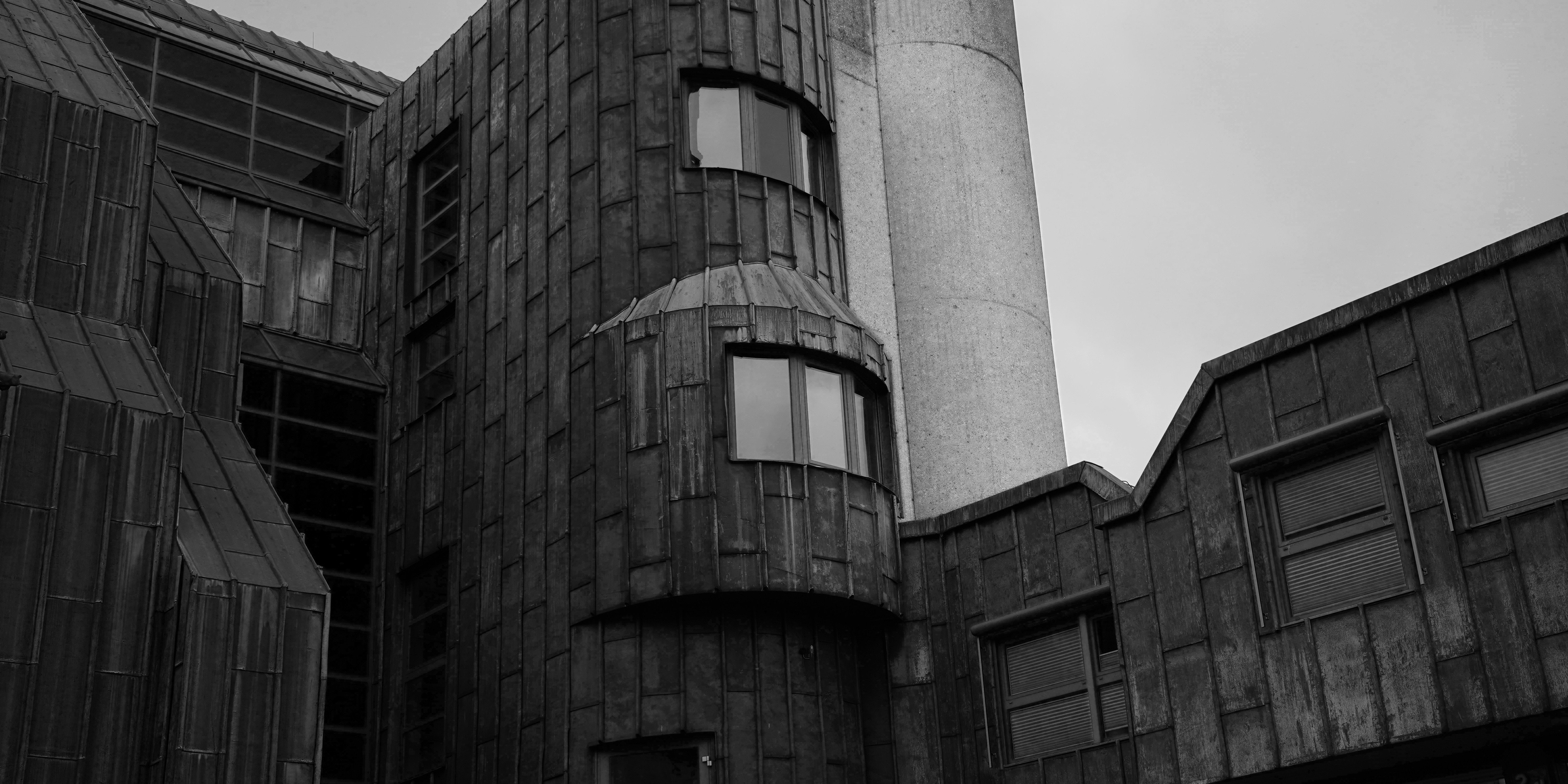 Monochrome exterior of brutalist industrial buildings with textured concrete, geometric windows, and a towering chimney, conveying stark urban massing.