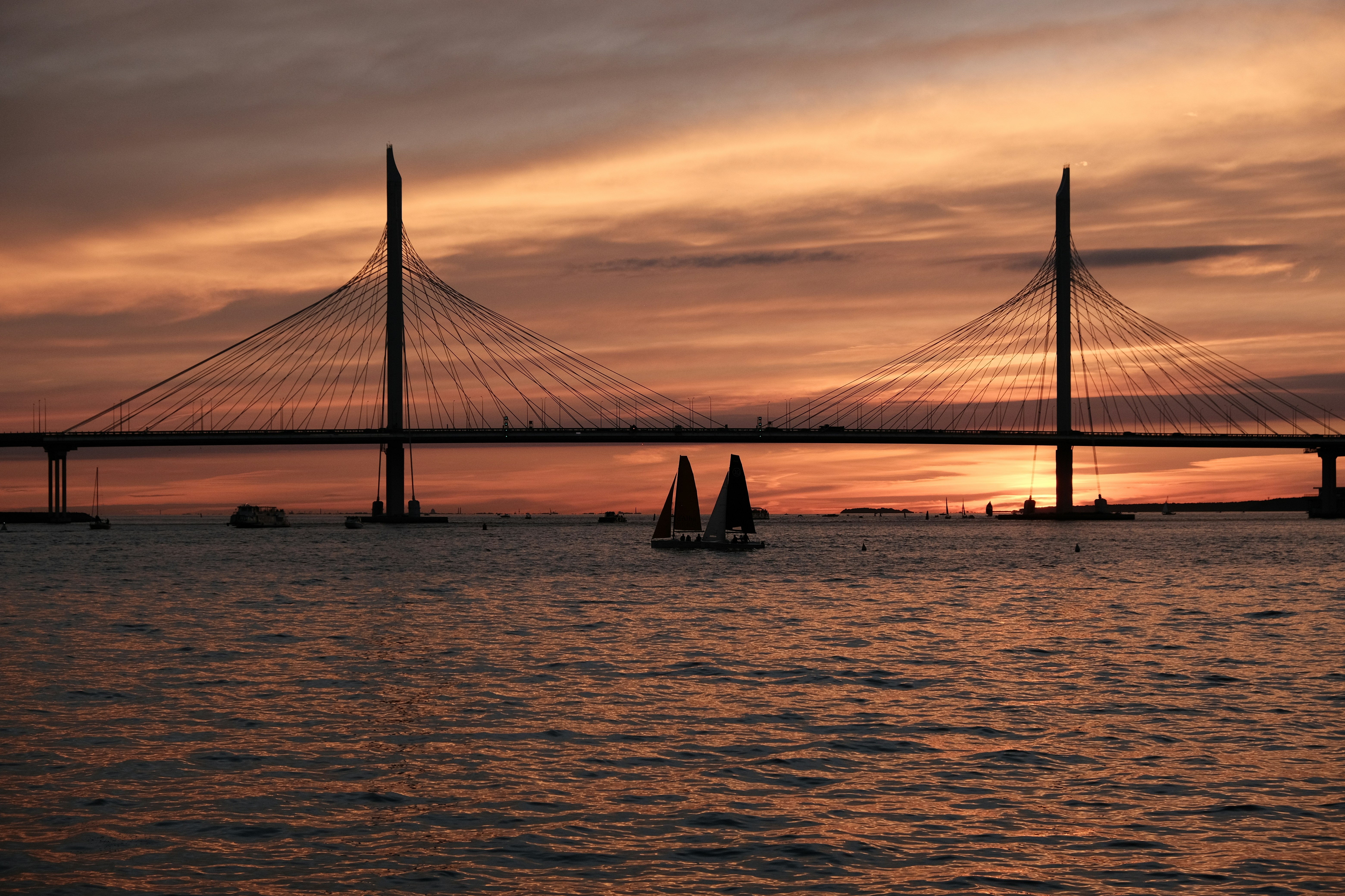 Silhouetted cable-stayed bridge at dusk spans a quiet body of water, with sailboats in the foreground and warm orange hues washing the horizon.