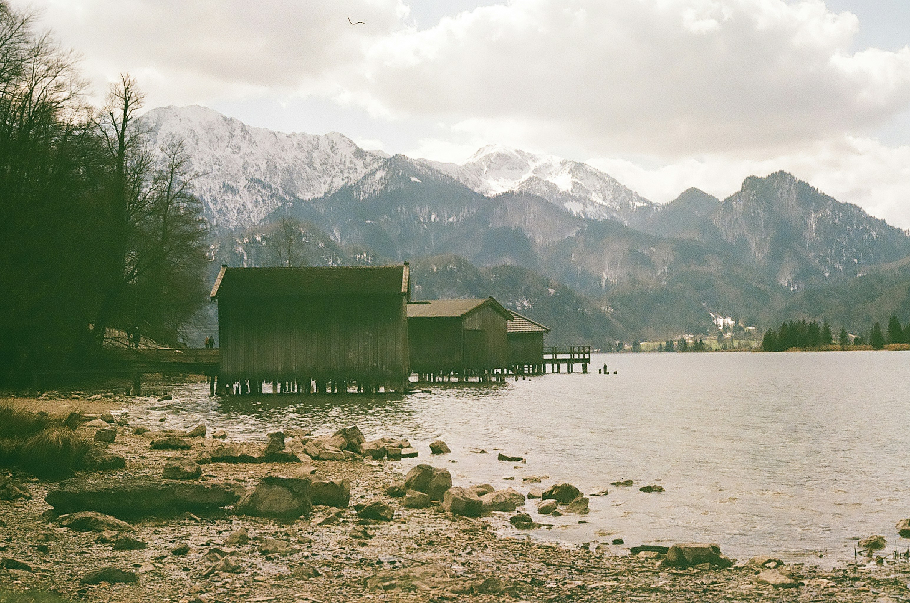Small wooden lakeside cabins extend over calm water with a rocky shore; distant snow-capped mountains provide a dramatic backdrop.