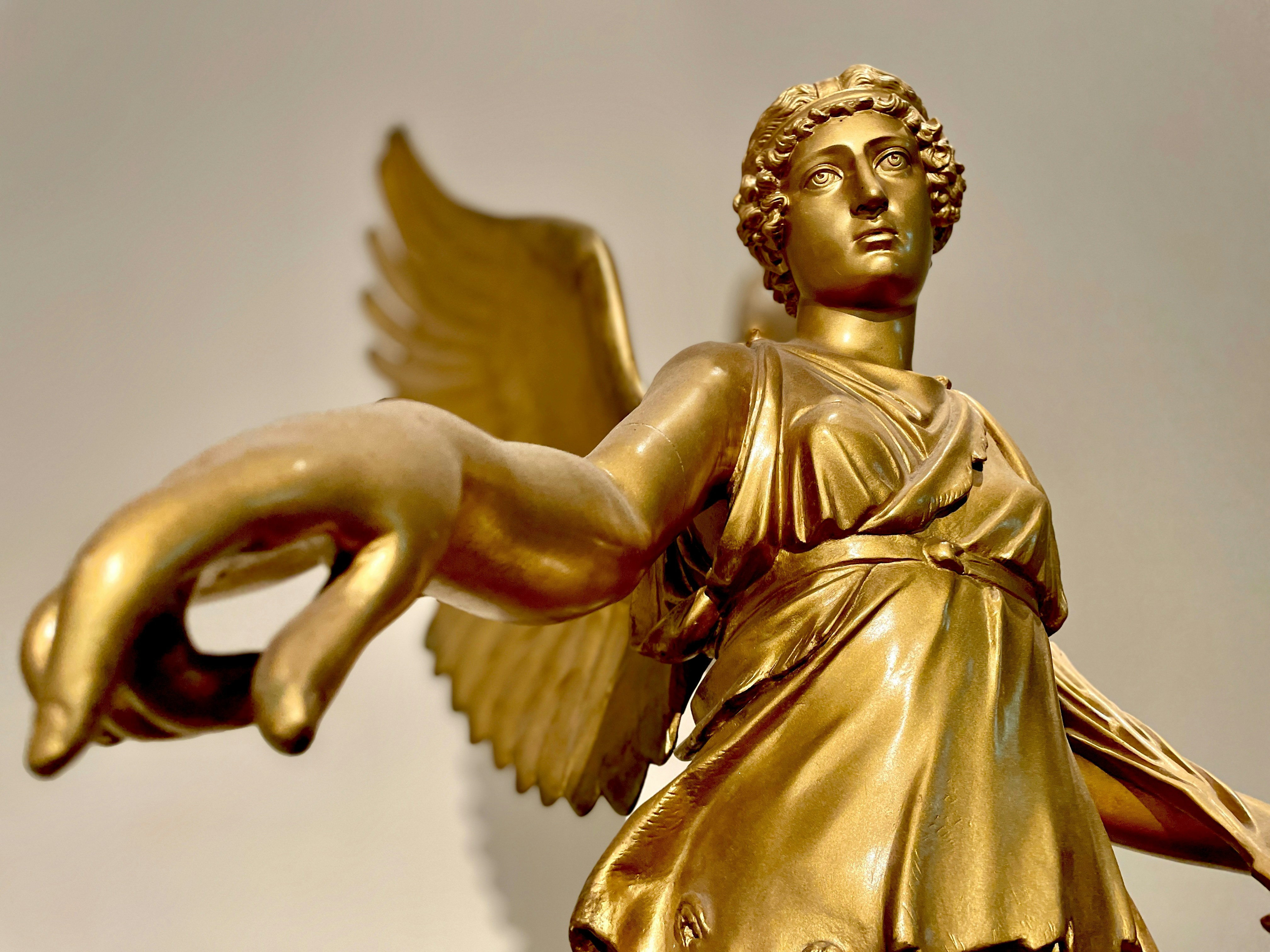 Golden angel statue with wing extended, dramatic light highlighting textured draped robes and sculpted facial features.