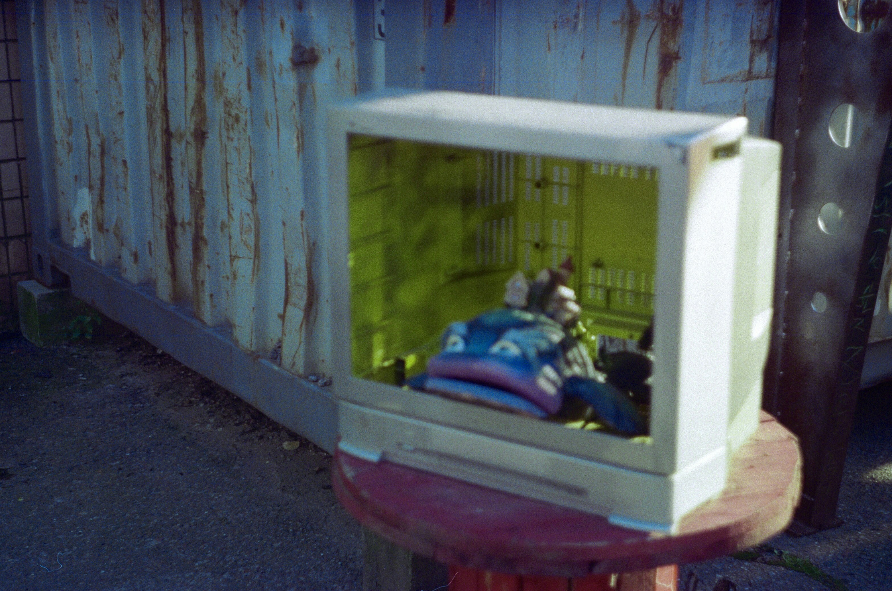 An old CRT television sits on a weathered red-painted table outdoors, its screen displaying a surreal image of a large, stylized fish with exaggerated features. The TV’s silver casing shows signs of age, and the screen’s glow casts a yellowish tint over the scene. Behind the set, a dilapidated metal container with peeling paint and rust streaks adds to the gritty, urban atmosphere. To the left, a section of black metal grid fencing is visible, while the ground is covered in rough asphalt. The lighting is cool and bluish, contrasting with the warm yellow of the screen and the red of the table.