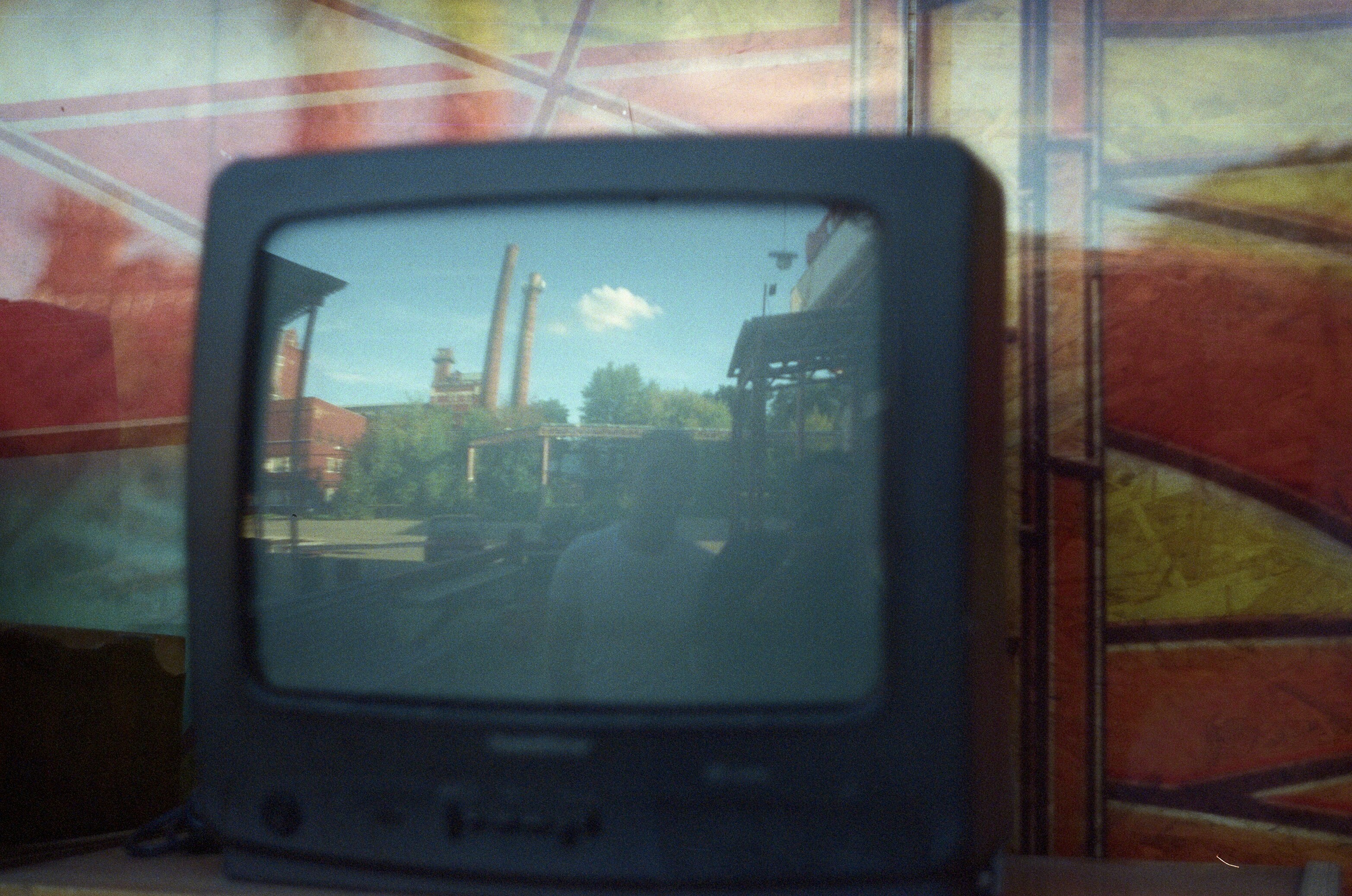 A vintage GoldStar CRT television sits in the foreground, its dark blue casing and physical control buttons evoking a sense of nostalgia for early 2000s electronics. The screen acts as a reflective surface, mirroring an industrial urban scene: twin factory chimneys rise against a clear blue sky, flanked by red-brick buildings and leafy green trees. In the reflection, the silhouettes of two people are faintly visible, adding a human element to the composition. The surrounding environment includes structural elements like a metal awning and street fixtures, subtly layered in the glass. The color