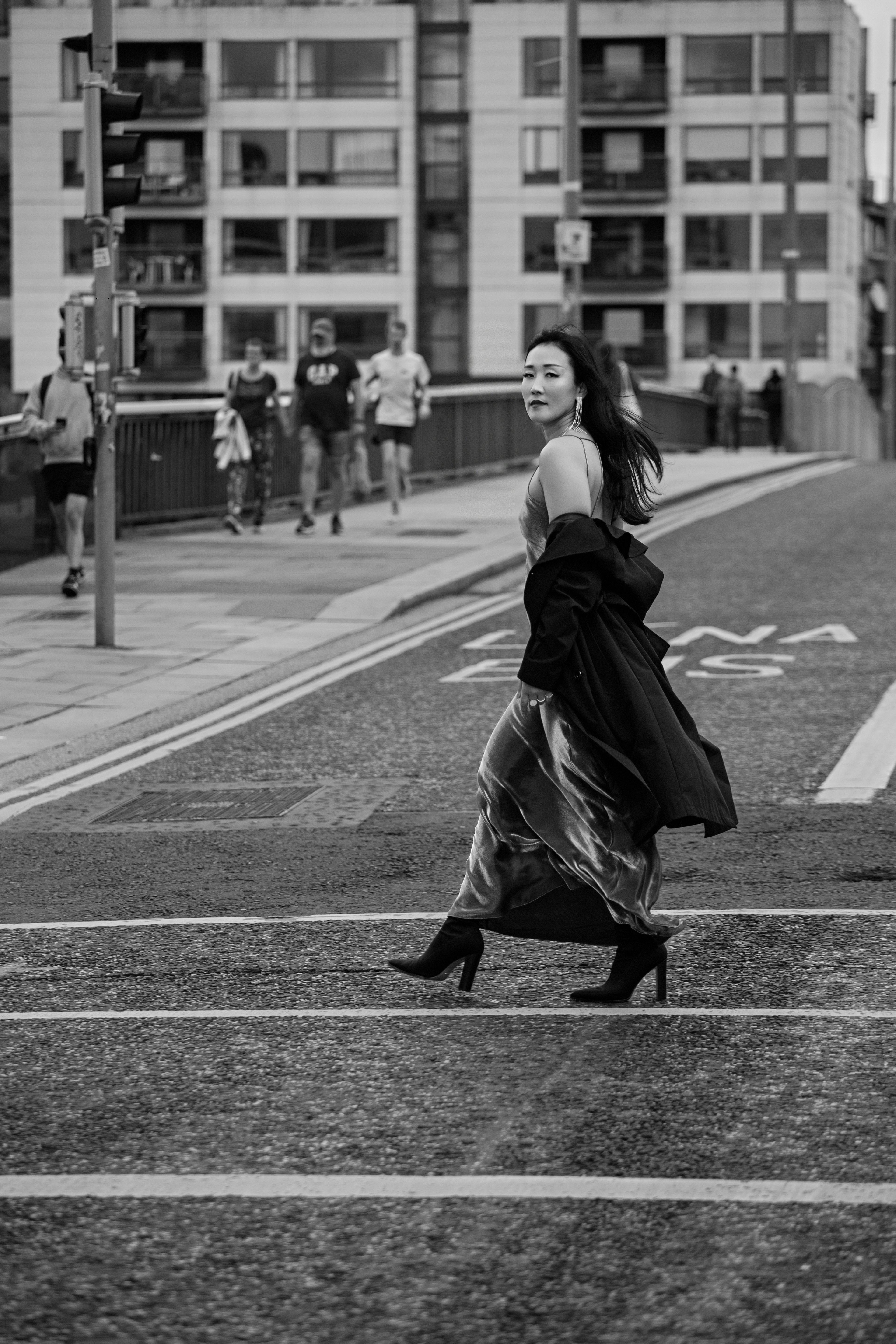 A stylish woman in a flowing dress and heels strides across a city street, jacket flowing behind her as pedestrians and modern buildings form a grayscale urban backdrop.