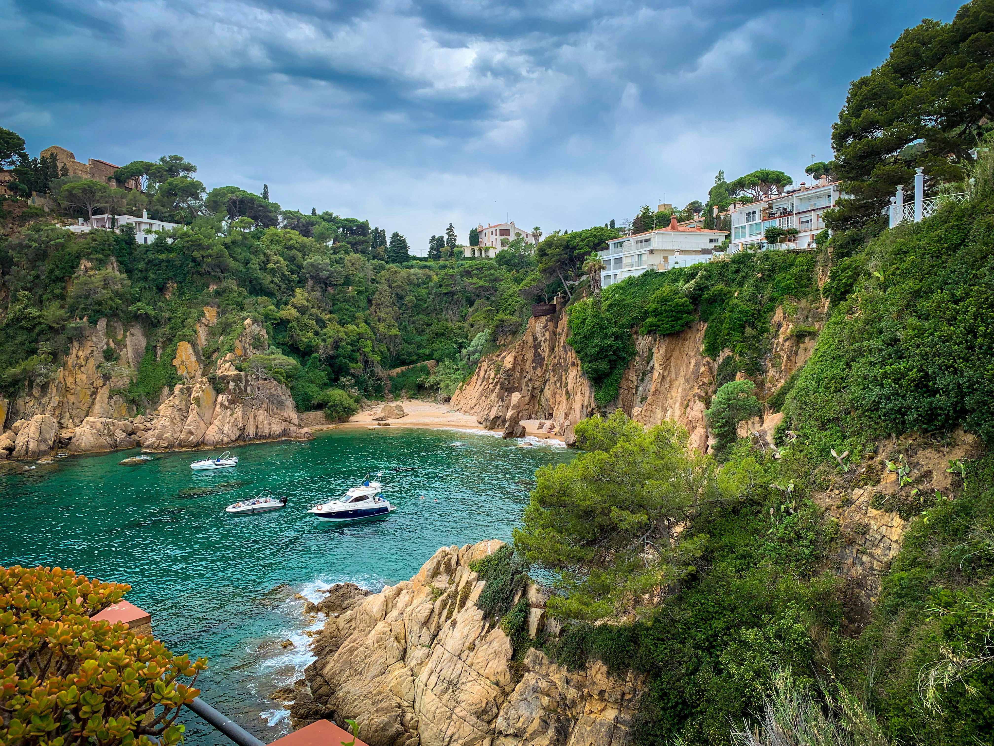 Vibrant coastal scene with a turquoise cove, rocky cliffs, and boats anchored near a sandy inlet; residential cliffside homes overlook the water.