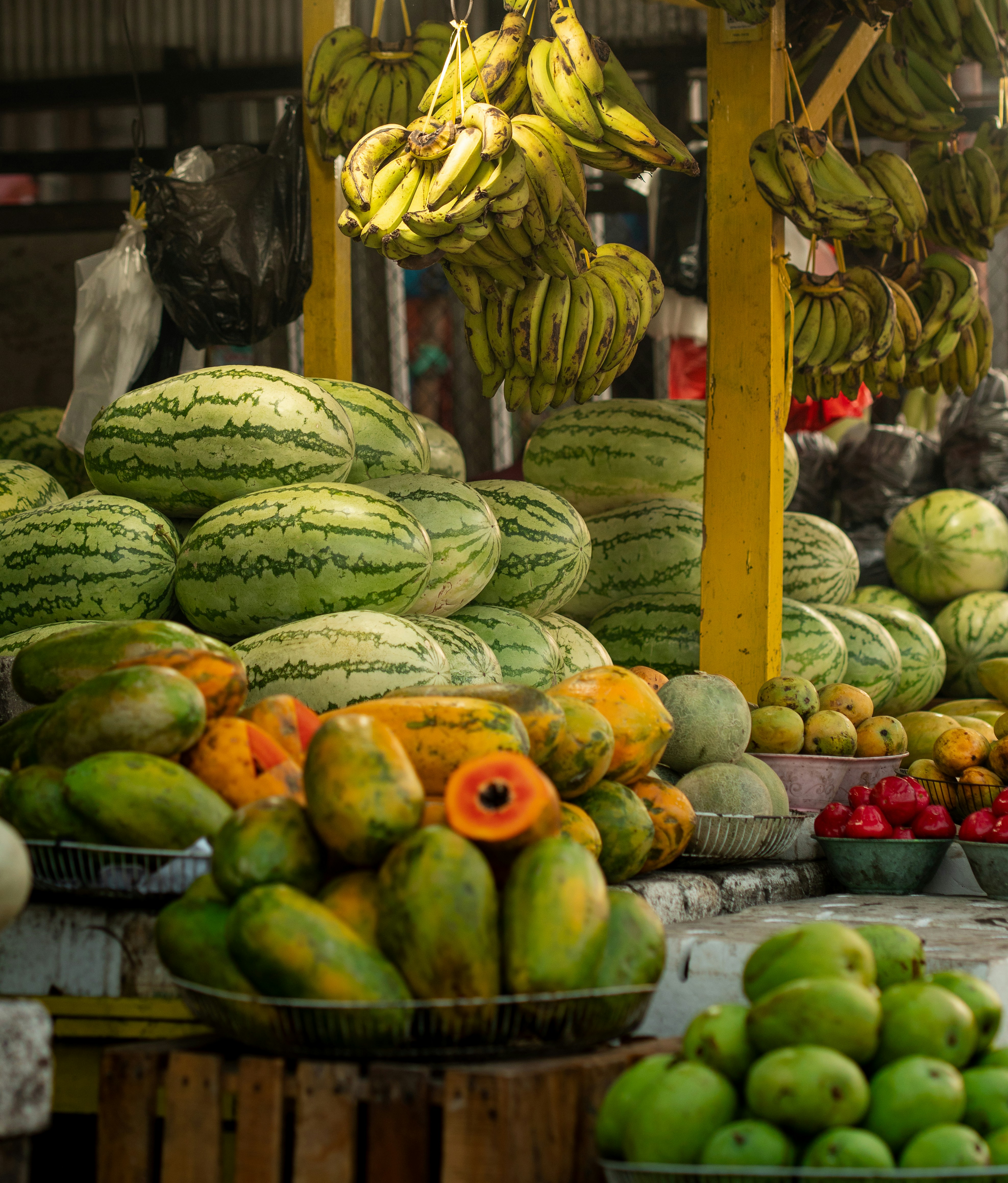 Vibrant display of watermelons and tropical fruit at a bustling market stall with hanging bananas overhead.