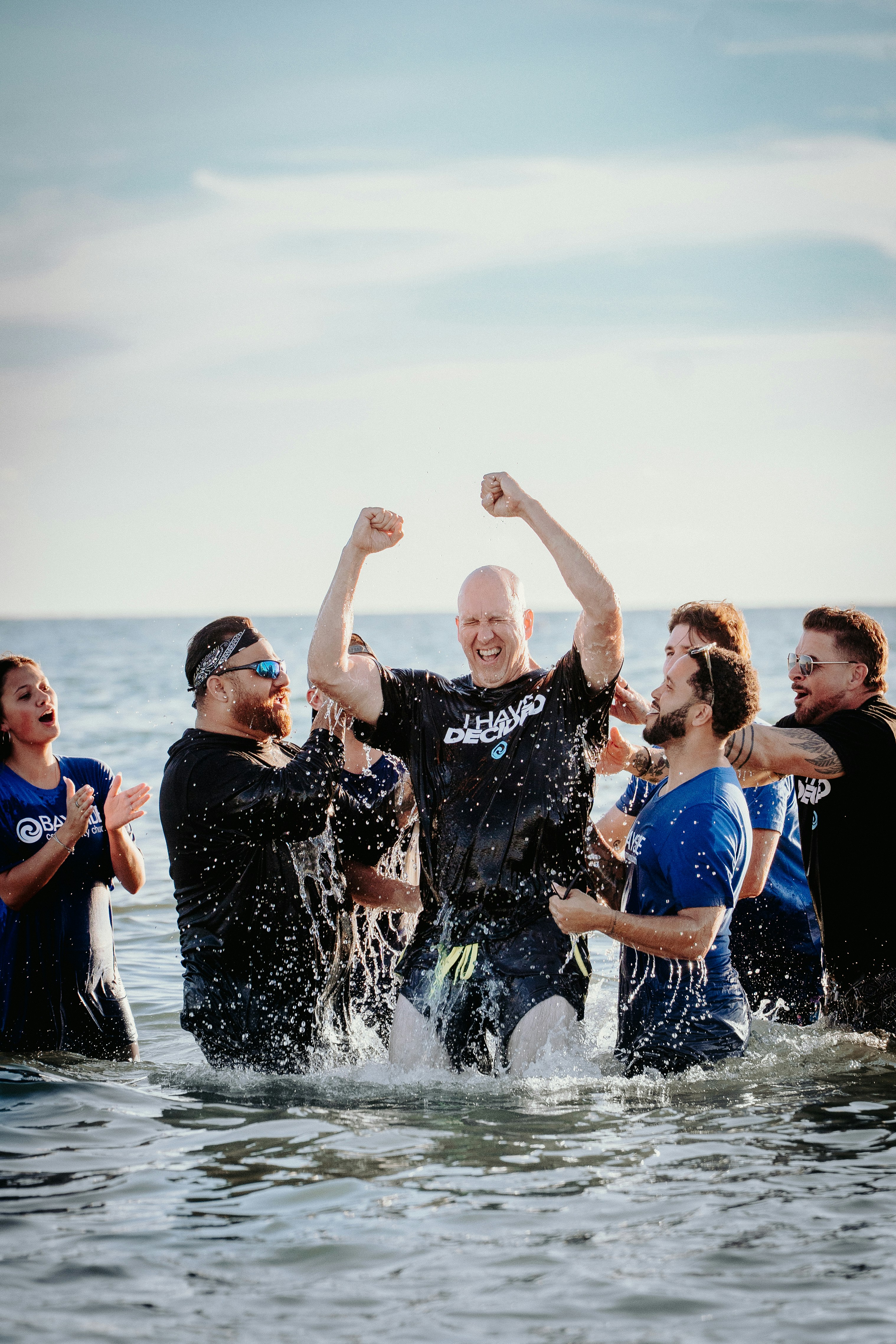 A group of people in ocean water celebrate with raised fists and smiles, suggesting a victory or team achievement; the scene focuses on the central bald man with others surrounding him, all in casual athletic wear.