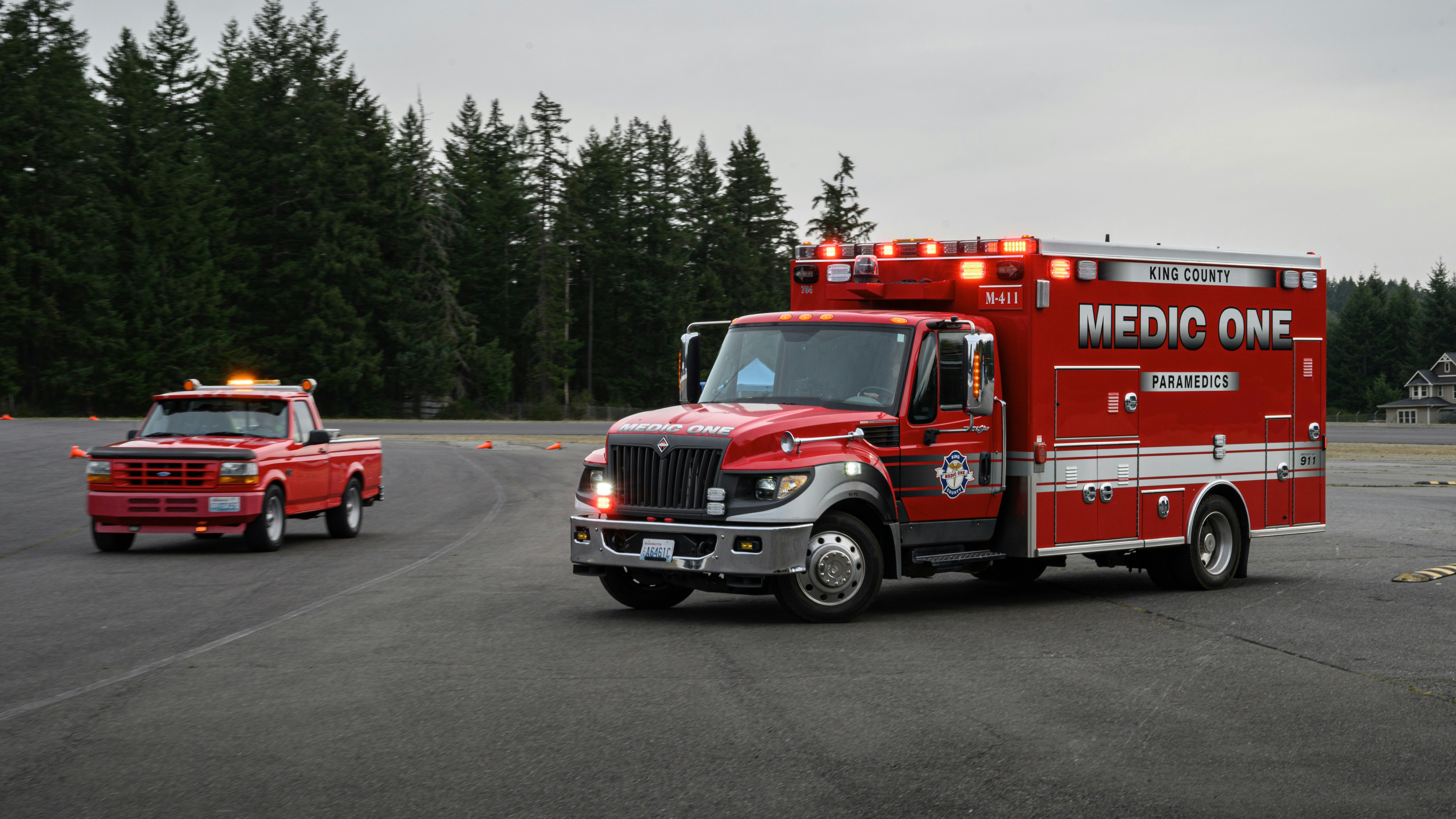 Red emergency ambulance leads a staggered convoy on an open tarmac, with a smaller red unit in the background and a forested line beyond, ready for response. Aimed at depicting readiness and coordination.