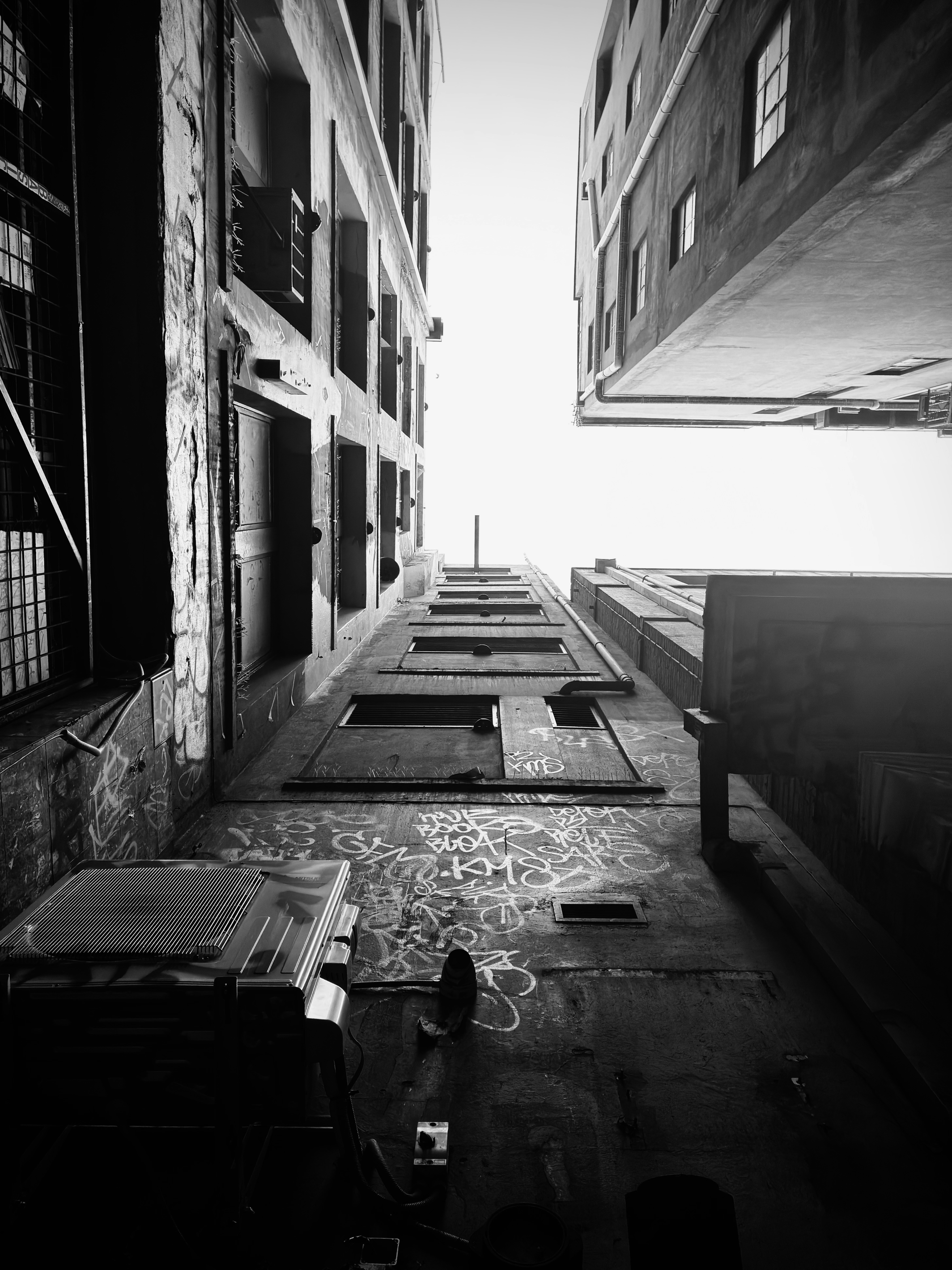 A black-and-white alleyway shot converges toward a bright opening above, with graffiti-covered ground and urban clutter filling the lower frame.