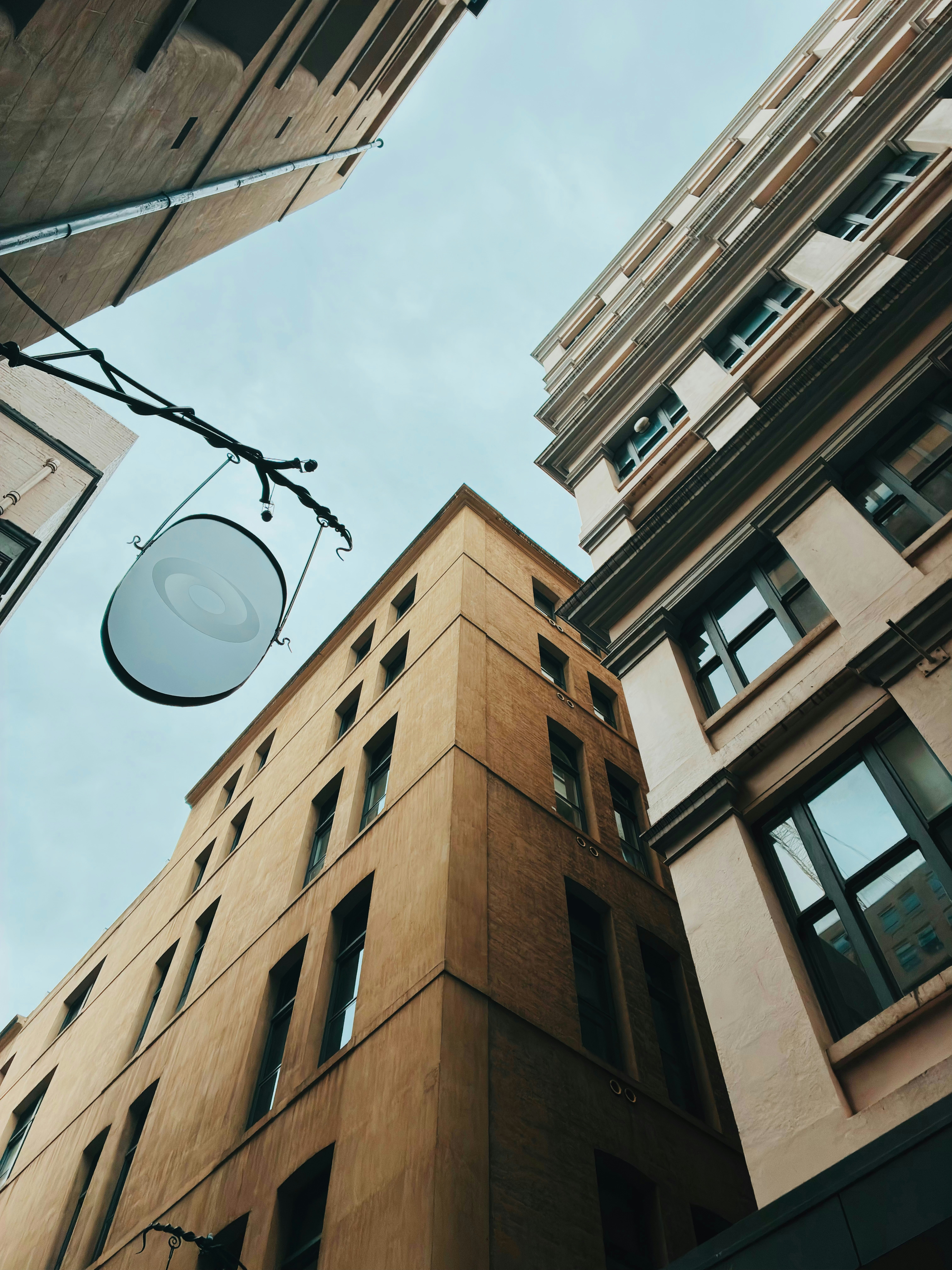 A tilted city scene with tall beige and white buildings framing a pale blue sky; a decorative circular streetlamp hangs in the foreground, adding a focal point to the urban composition.