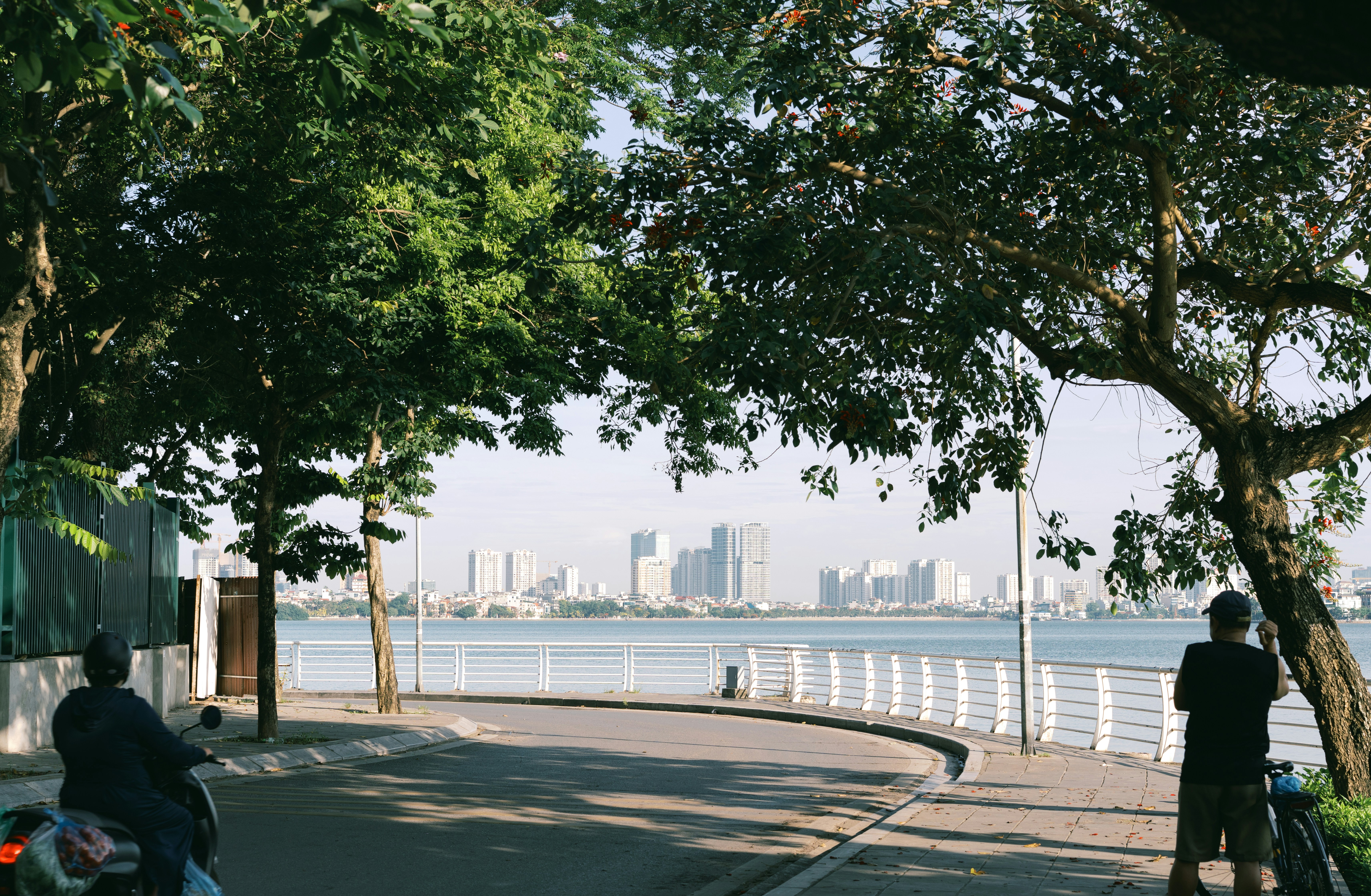 Riverside path with pedestrians under leafy branches, overlooking a skyline across the water.
