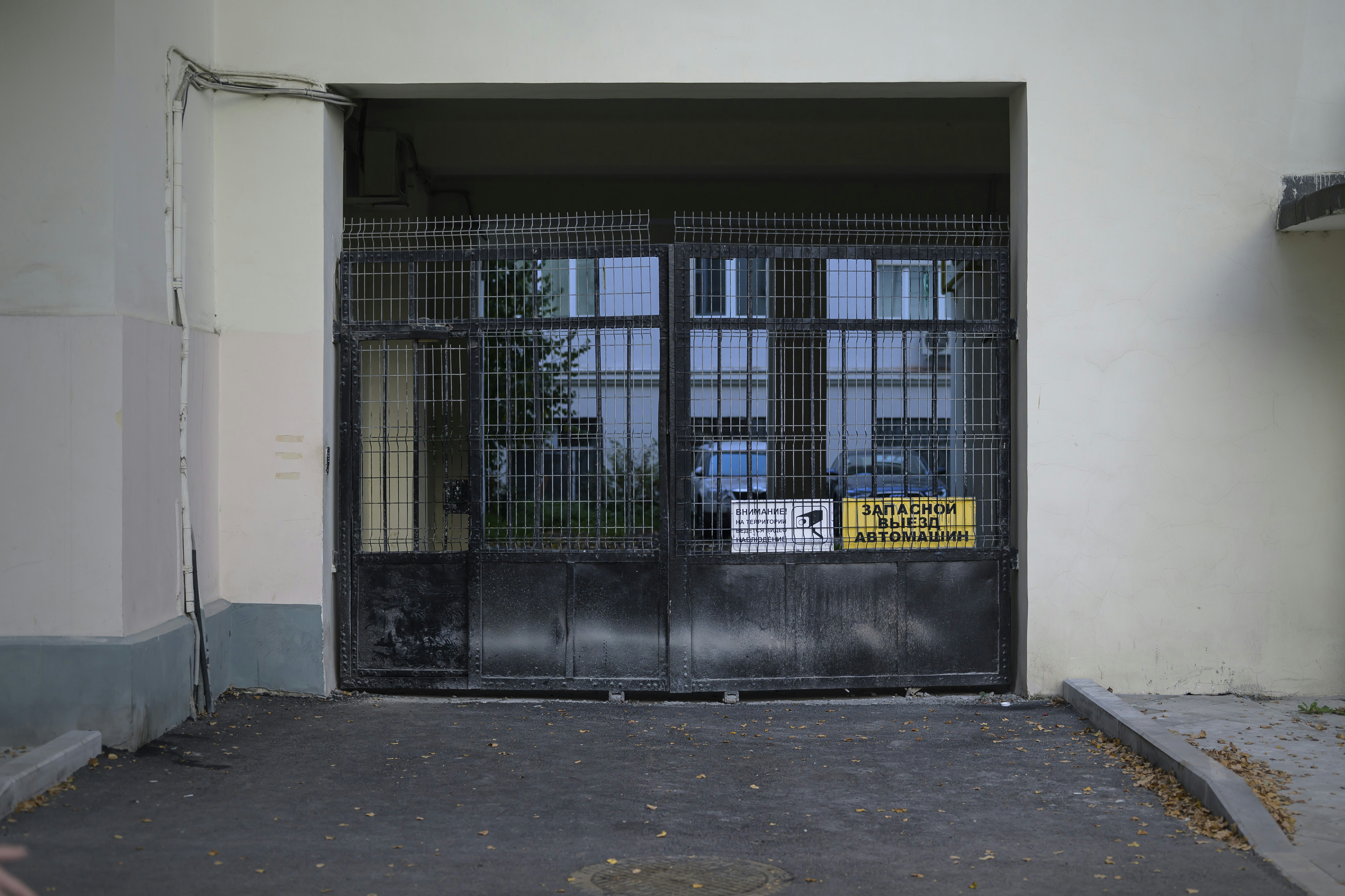 A quiet courtyard entrance in Yekaterinburg framed by a heavy metal gate and pale plaster walls. The black grille reveals a glimpse of parked cars and apartment windows beyond, while small warning signs, cracked paint and scattered autumn leaves on the pavement tell a story of everyday city life. Muted tones, soft natural light and balanced composition turn a utilitarian threshold into a calm, textured study of urban detail.