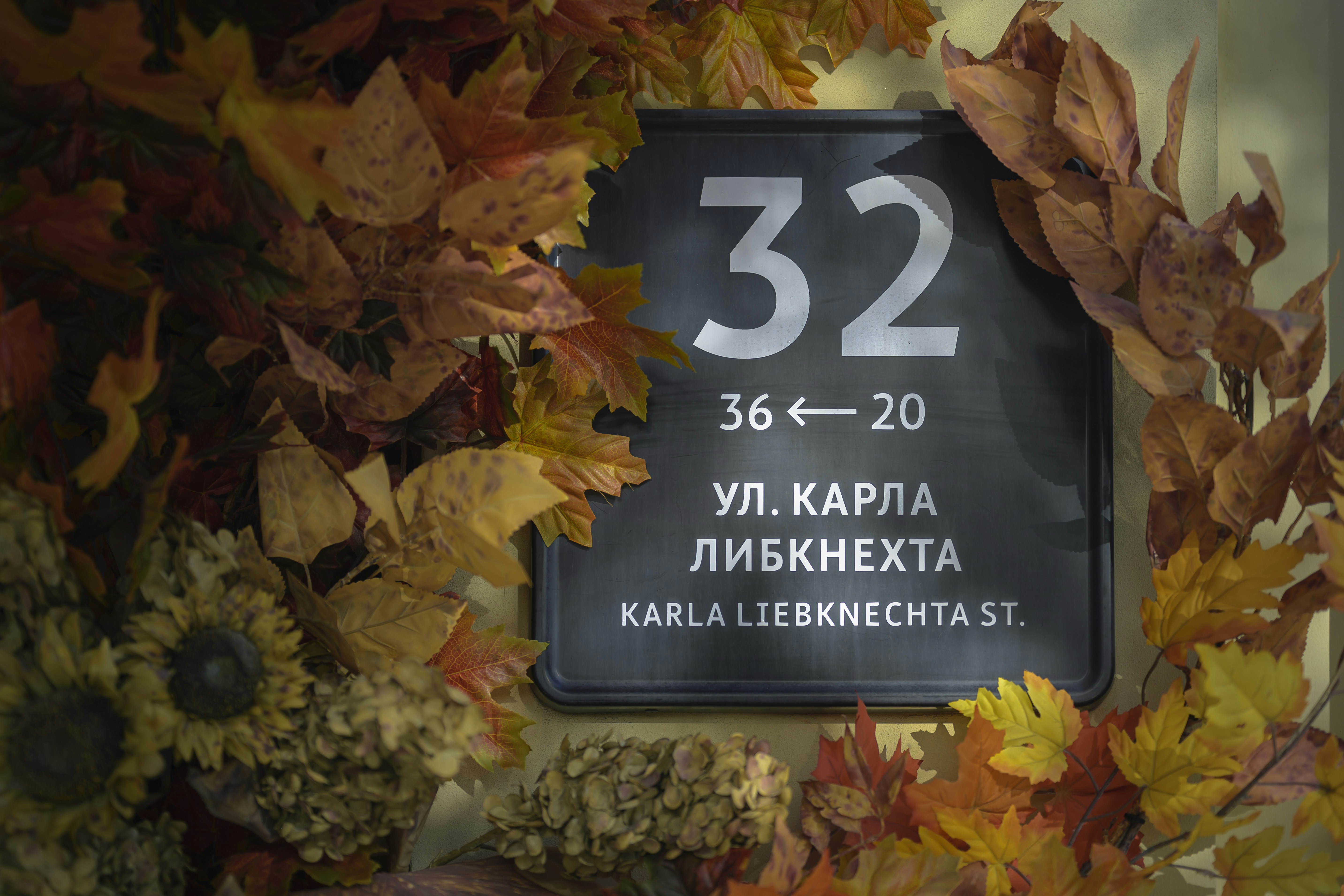 A matte black address plate for No. 32 on Karla Liebknechta Street in Yekaterinburg, framed by a halo of autumn leaves and dried blooms. Warm, directional light brings out the bold numerals and clean bilingual typography, contrasting the cool sign with rich seasonal tones. The close-up composition turns a simple building marker into a textured, intimate still life of city detail and fall atmosphere.