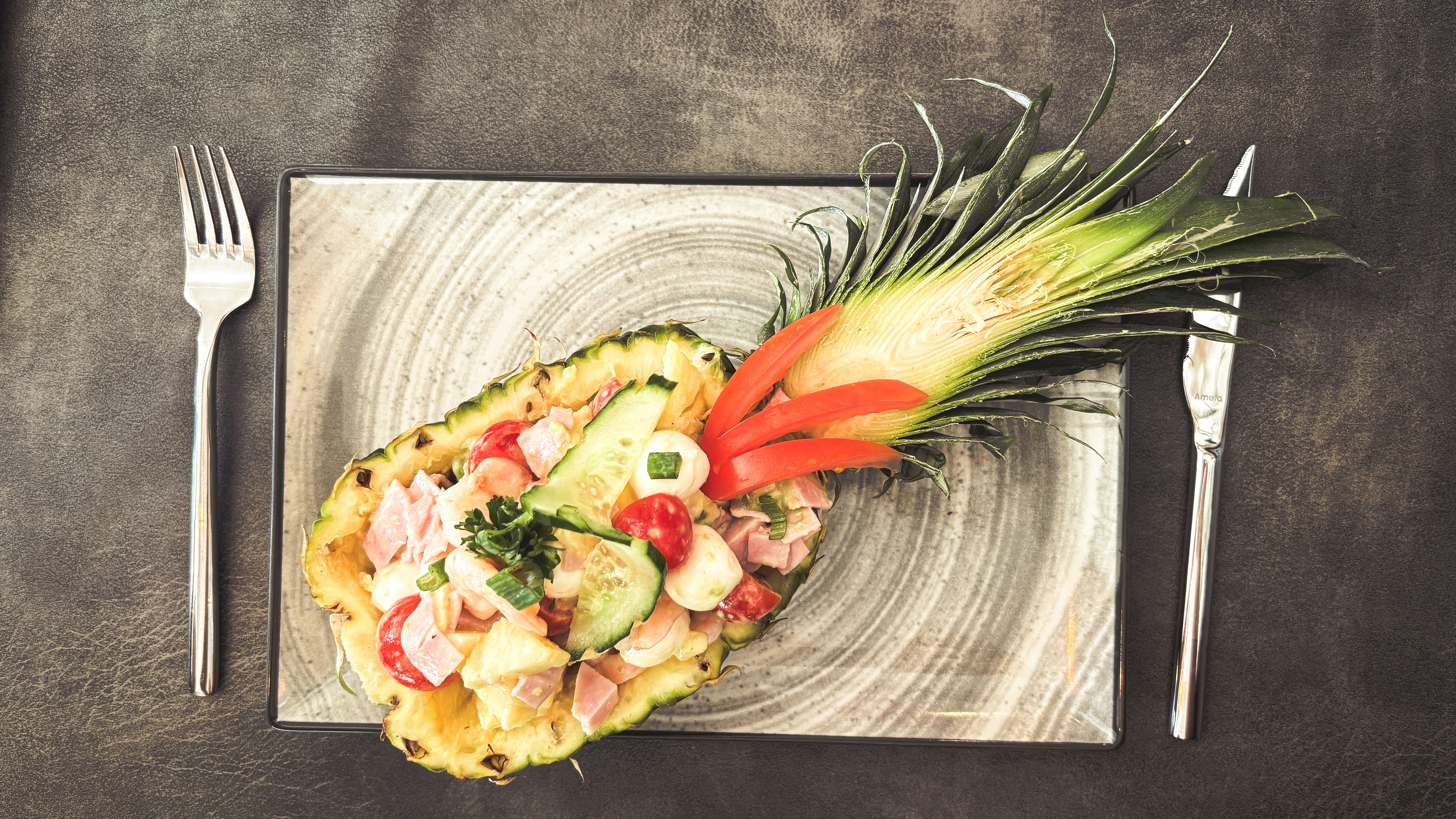 Savory pineapple boat filled with chunks of ham, tomato, onion and herbs presented on a rustic plate. A playful tropical appetizer highlighting bright colors and fresh ingredients.