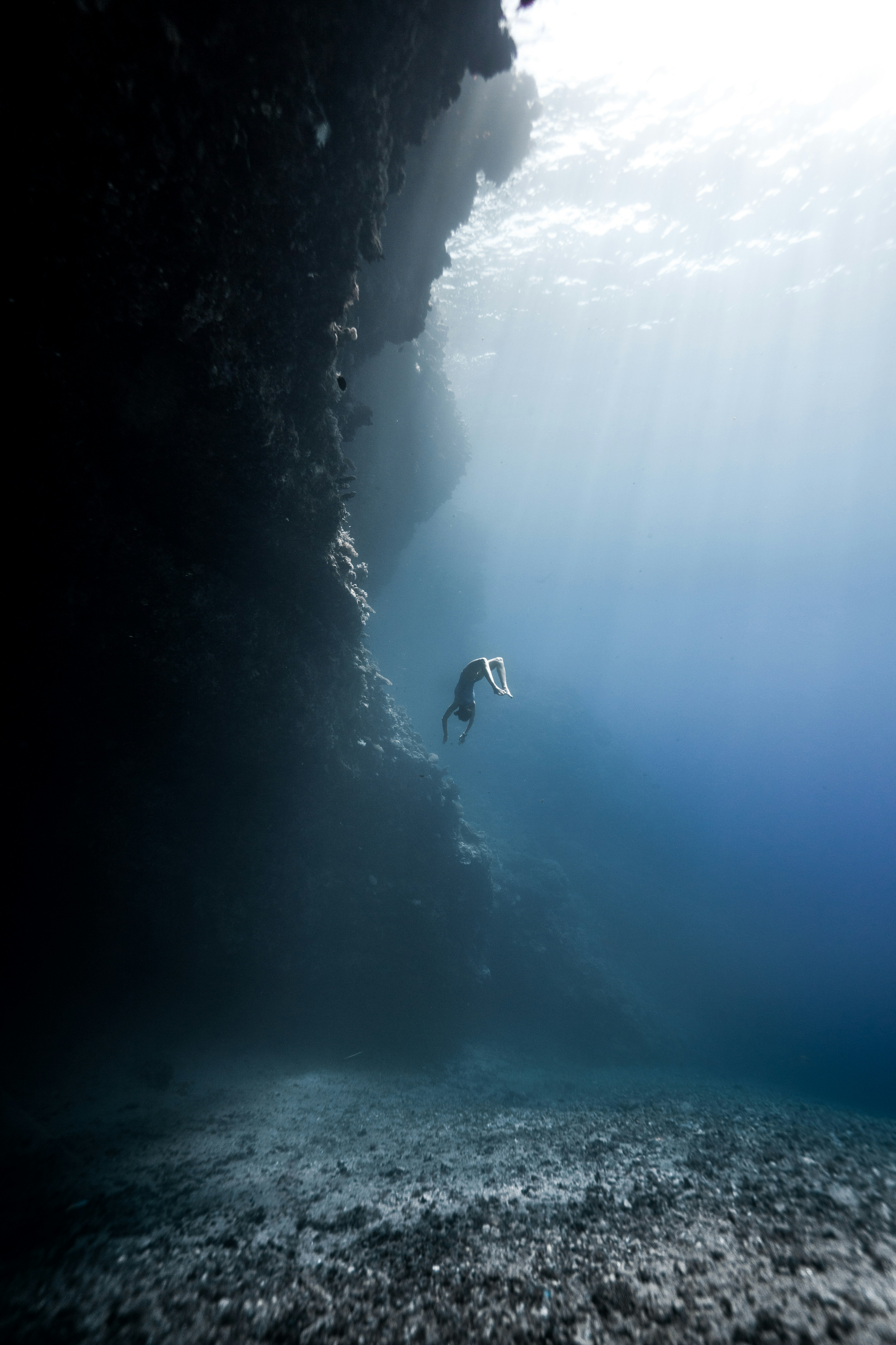 A lone diver glides along a sunlit underwater canyon, with rocky walls and a sandy seabed, bathed in blue light. This is a natural underwater photograph.