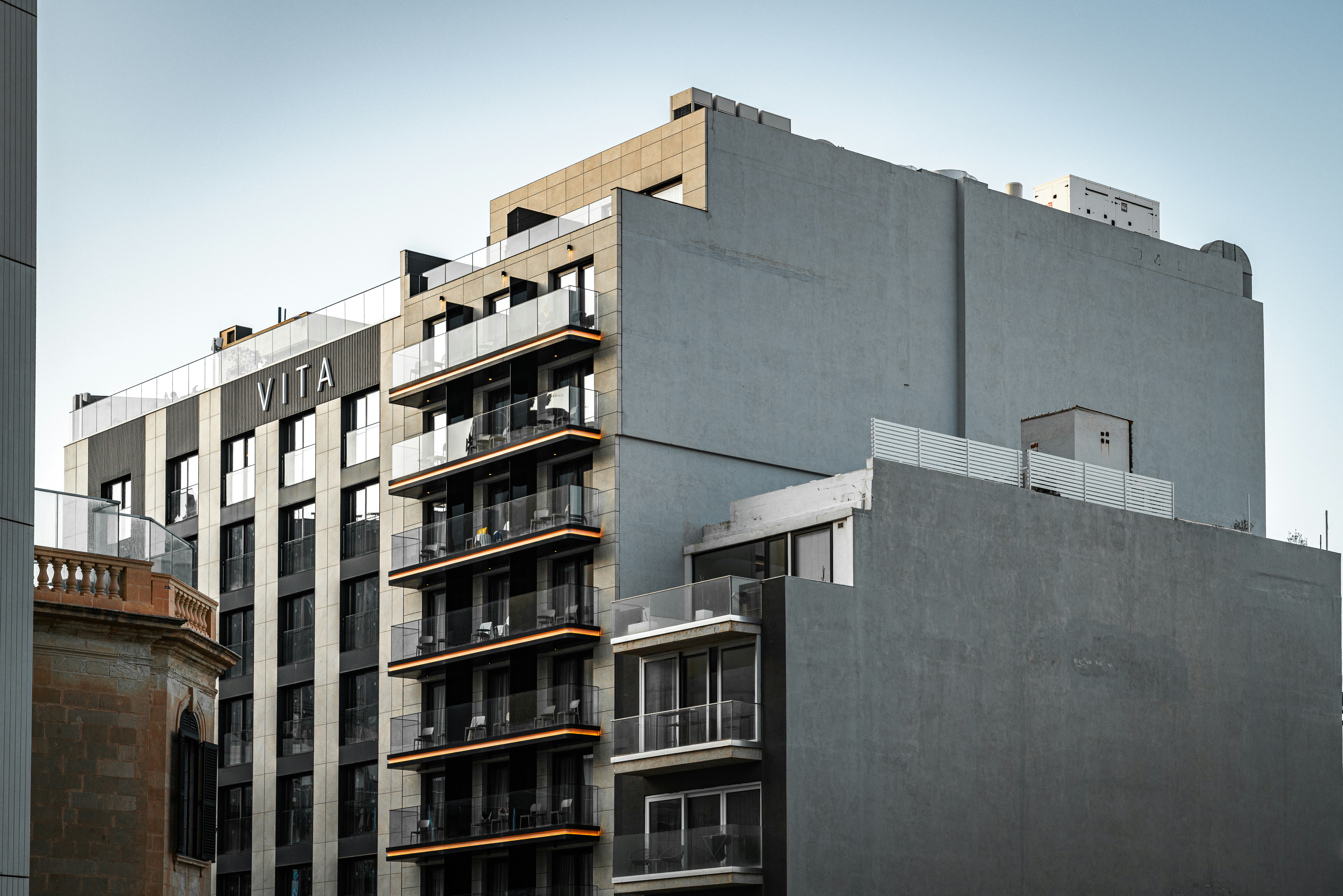 Tall, geometric apartment blocks rise against a pale sky, with a mix of glass and concrete textures and a hint of warm balcony railings.