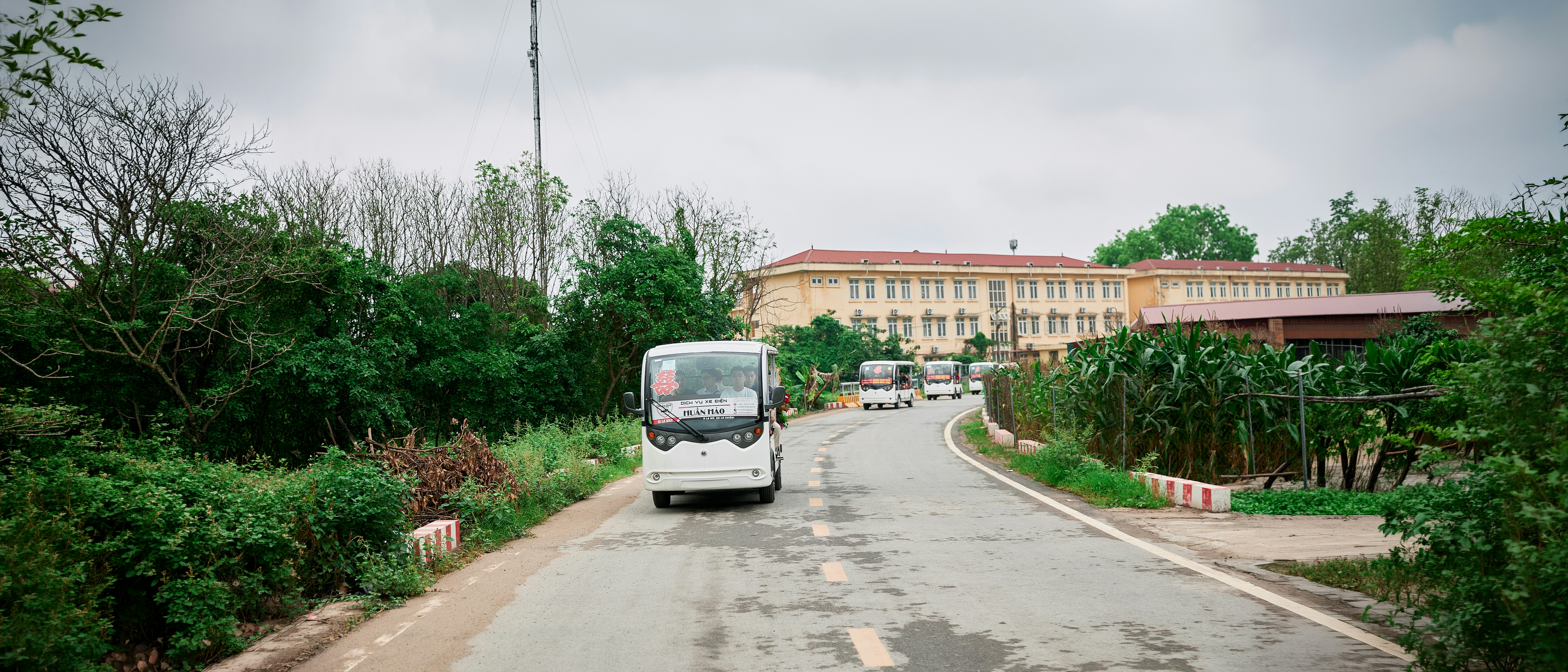 Concrete road leads toward a yellow building with greenery on both sides. A small white bus travels the centerline, suggesting a campus or suburban complex.