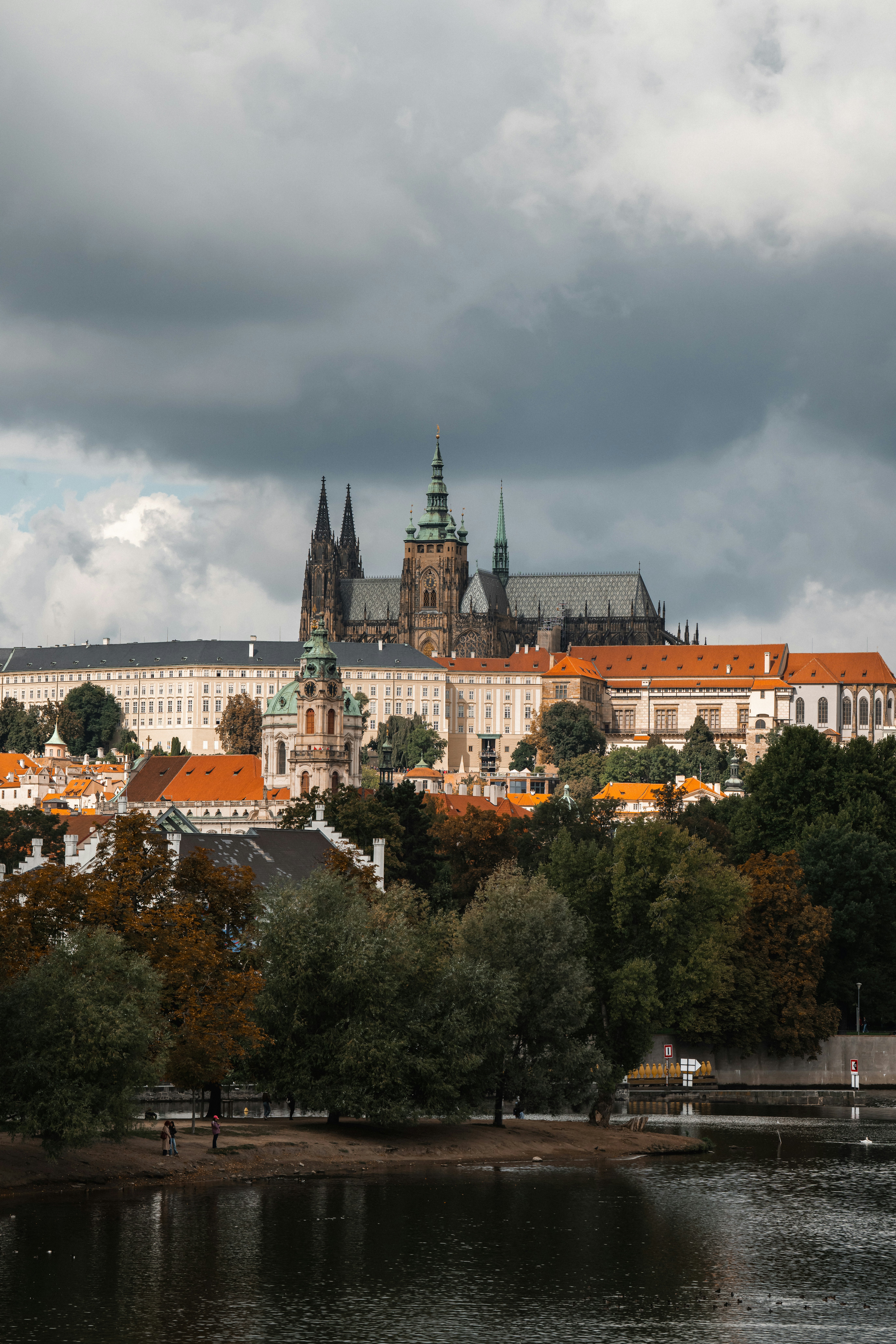 Autumnal cityscape with historic cathedral spires rising above red-tiled roofs along a riverbank; a calm water foreground and a dramatic cloud-filled sky frame the scene.