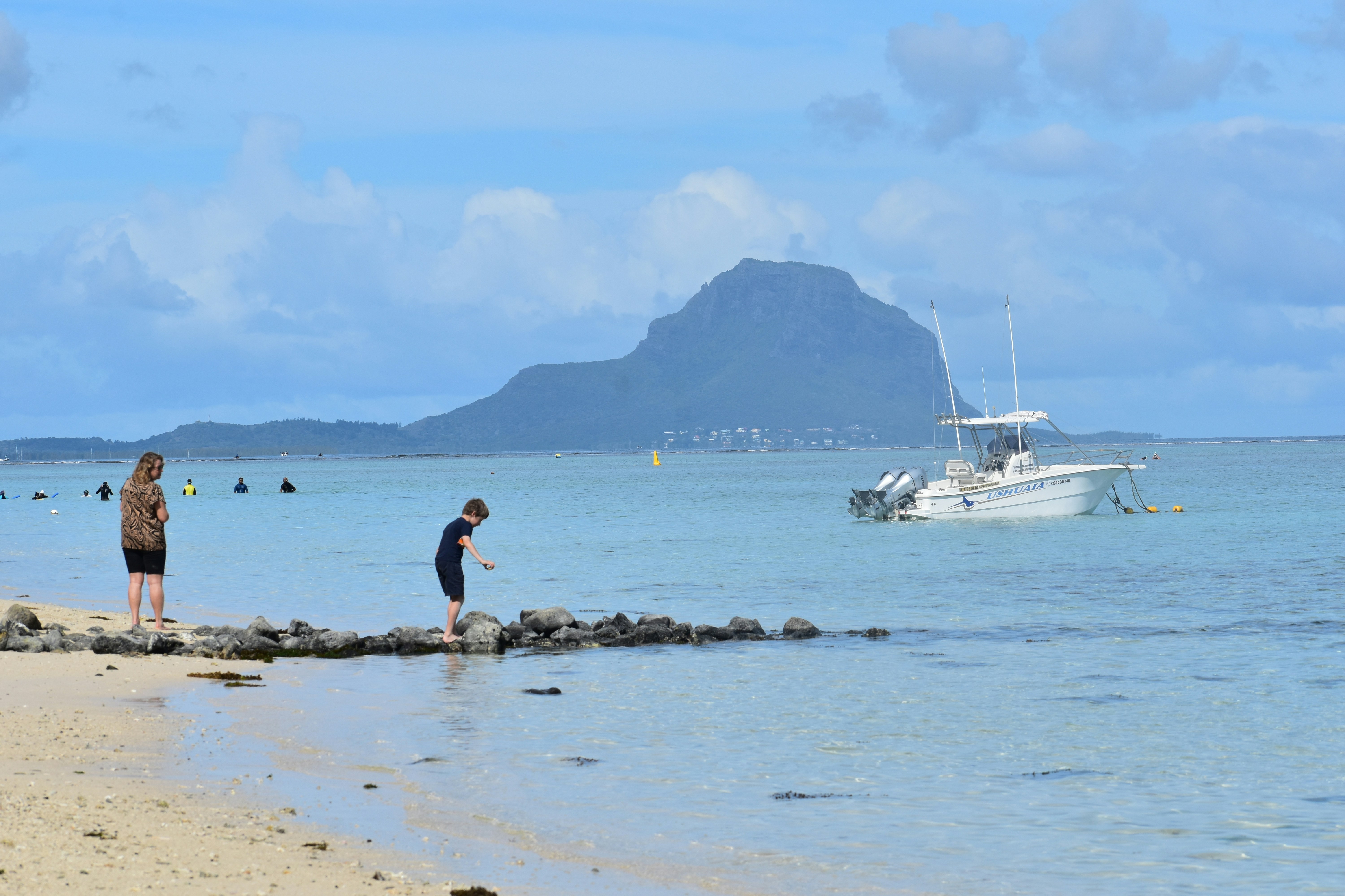 Beachgoers on a rocky jetty with a distant sailboat and island silhouette