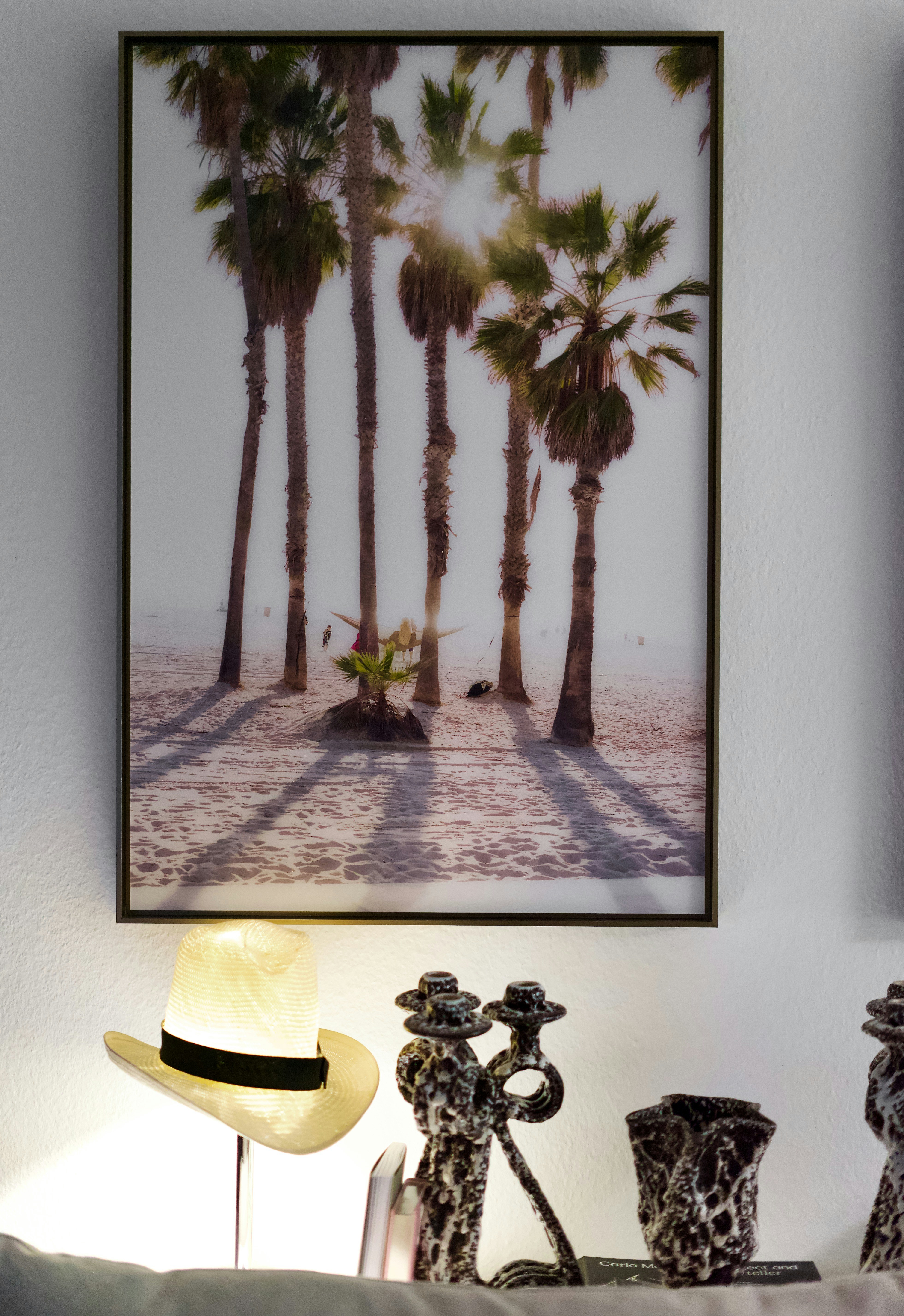 Venice Beach Palms & Warm Interior Vignette. A framed fine art print of California palm trees hanging in a warm, curated interior alongside a lit hat lamp and mid-century ceramics.
