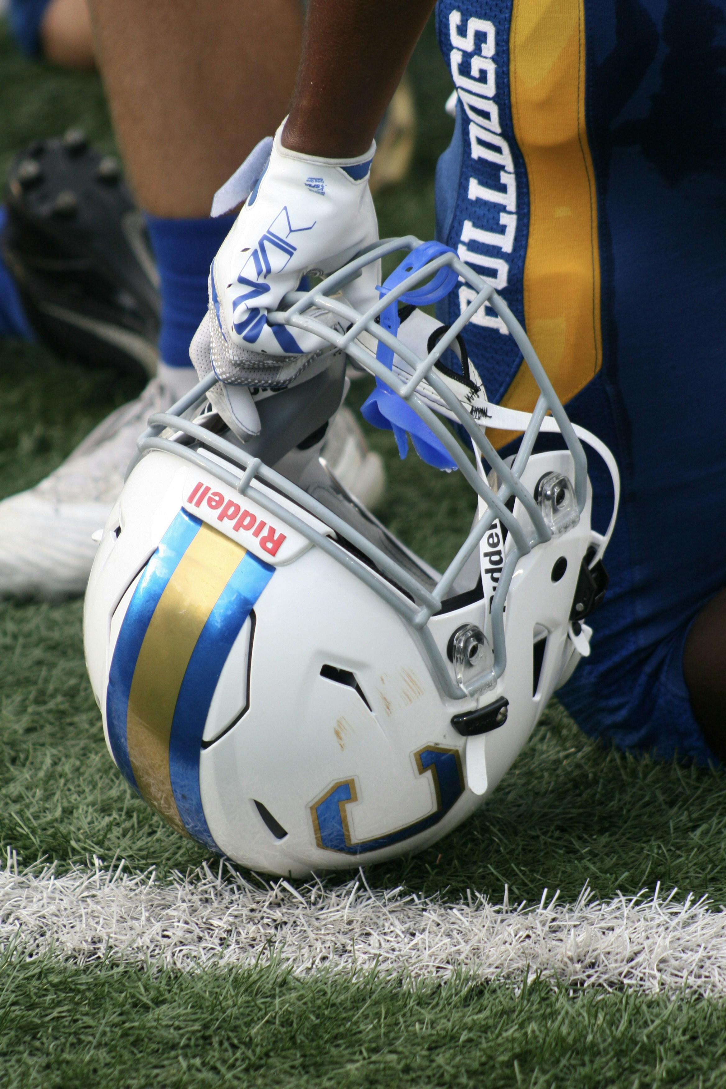 Football helmet resting on grass with cleats and uniform visible; close composition emphasizes team colors and wearables, conveying a moment of pause or preparation. Logo and gear are clear, indicating athletic context.