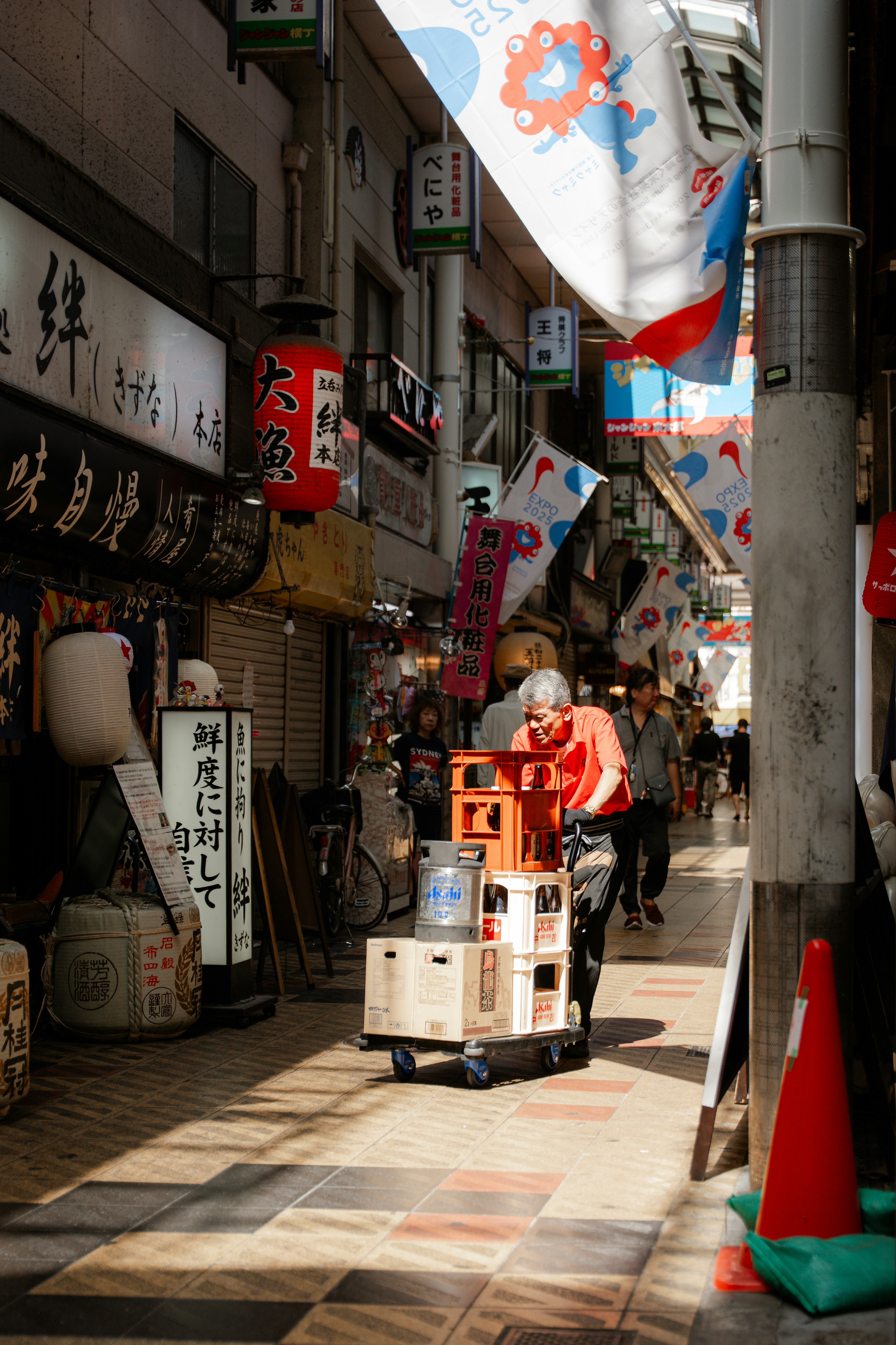 A man pushing a loaded cart down a narrow, sunlit street lined with shopfronts and hanging banners.