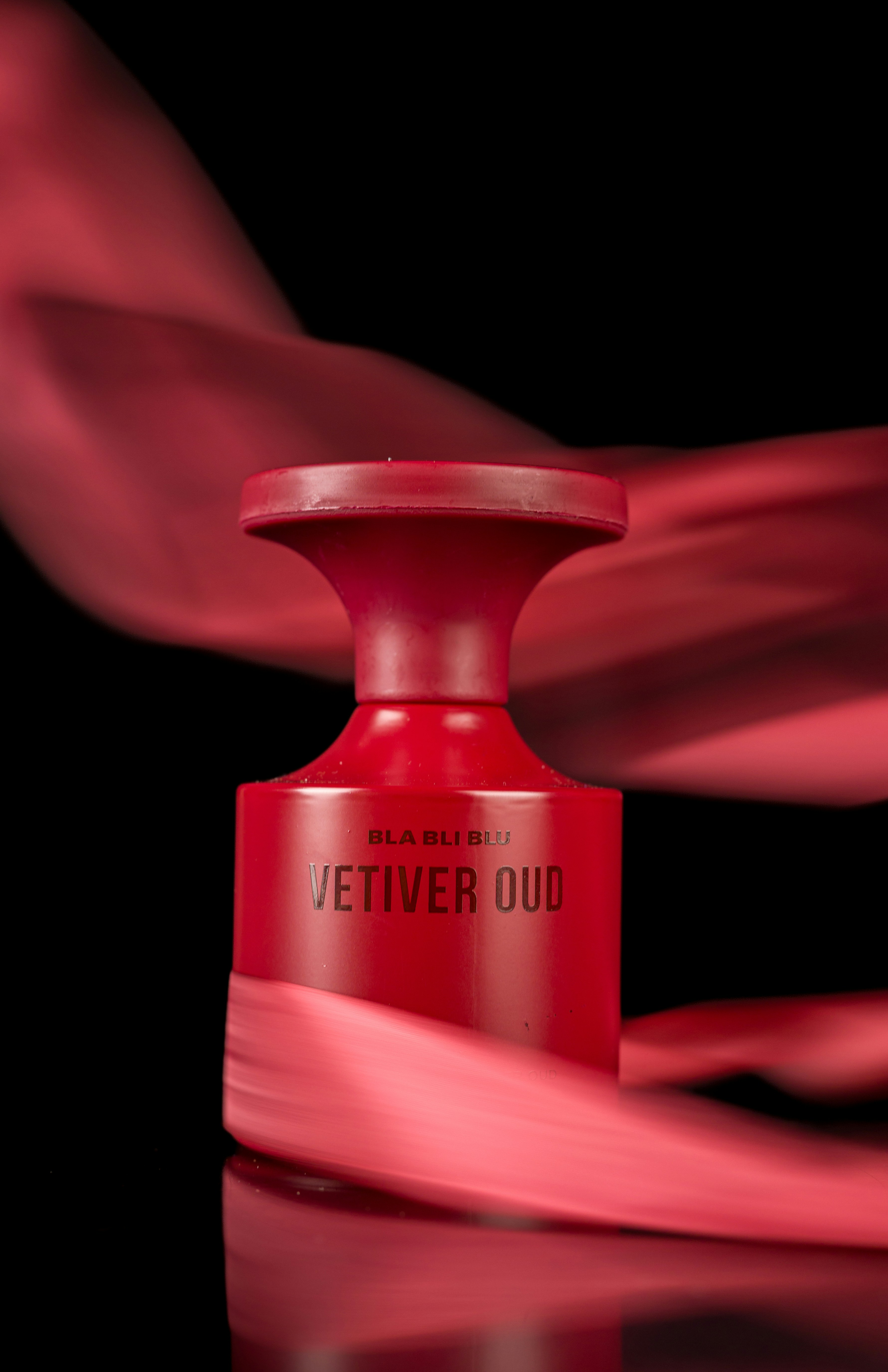 Red perfume bottle with flowing fabric-like ribbons swirling around it against a black background. The composition centers on the bottle’s cap and branding, conveying motion and elegance. This is a product shot.