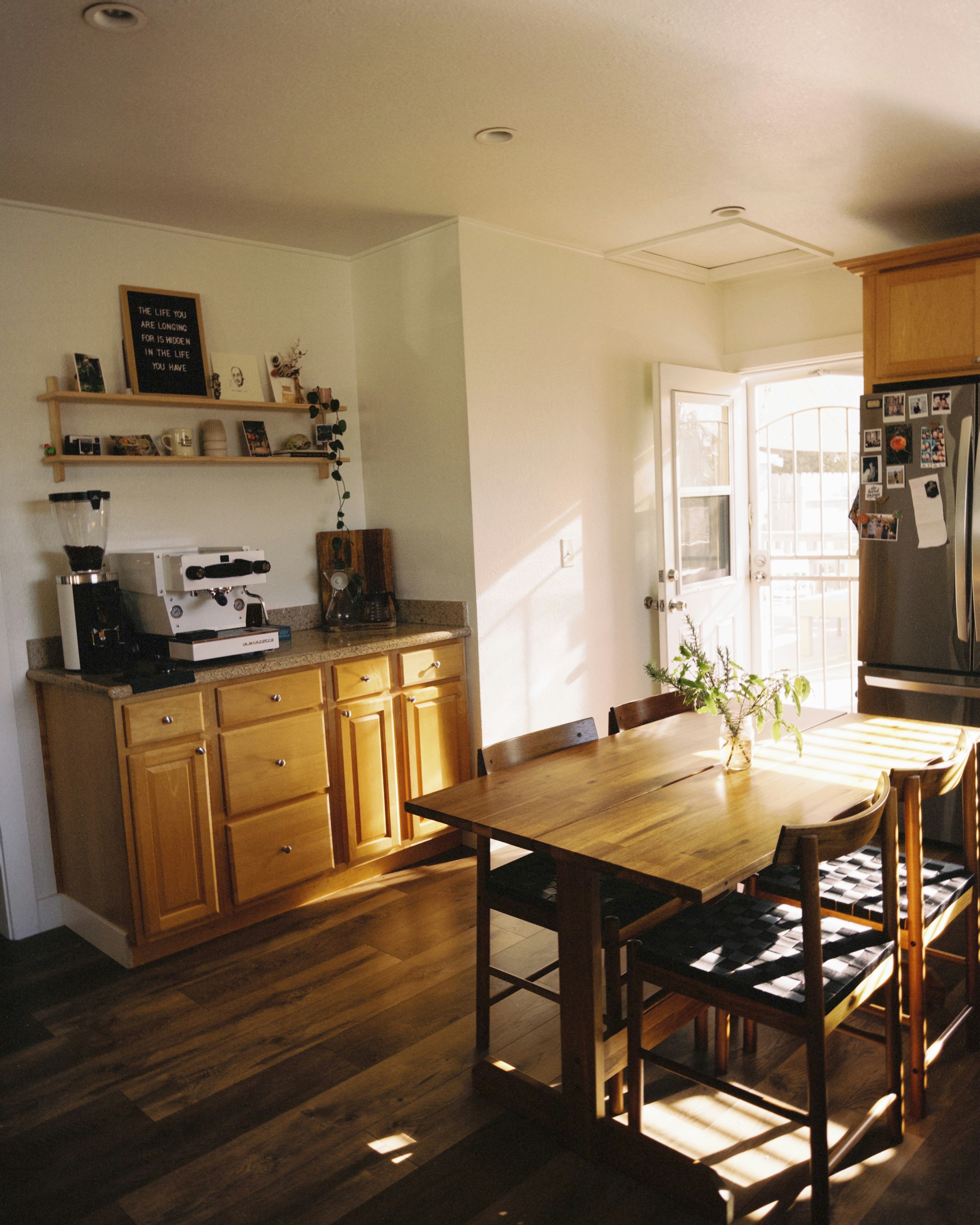 Sunlit dining area in a warm kitchen featuring a wooden table set for four, a coffee station along the left counter, and light streaming through a glass door.
