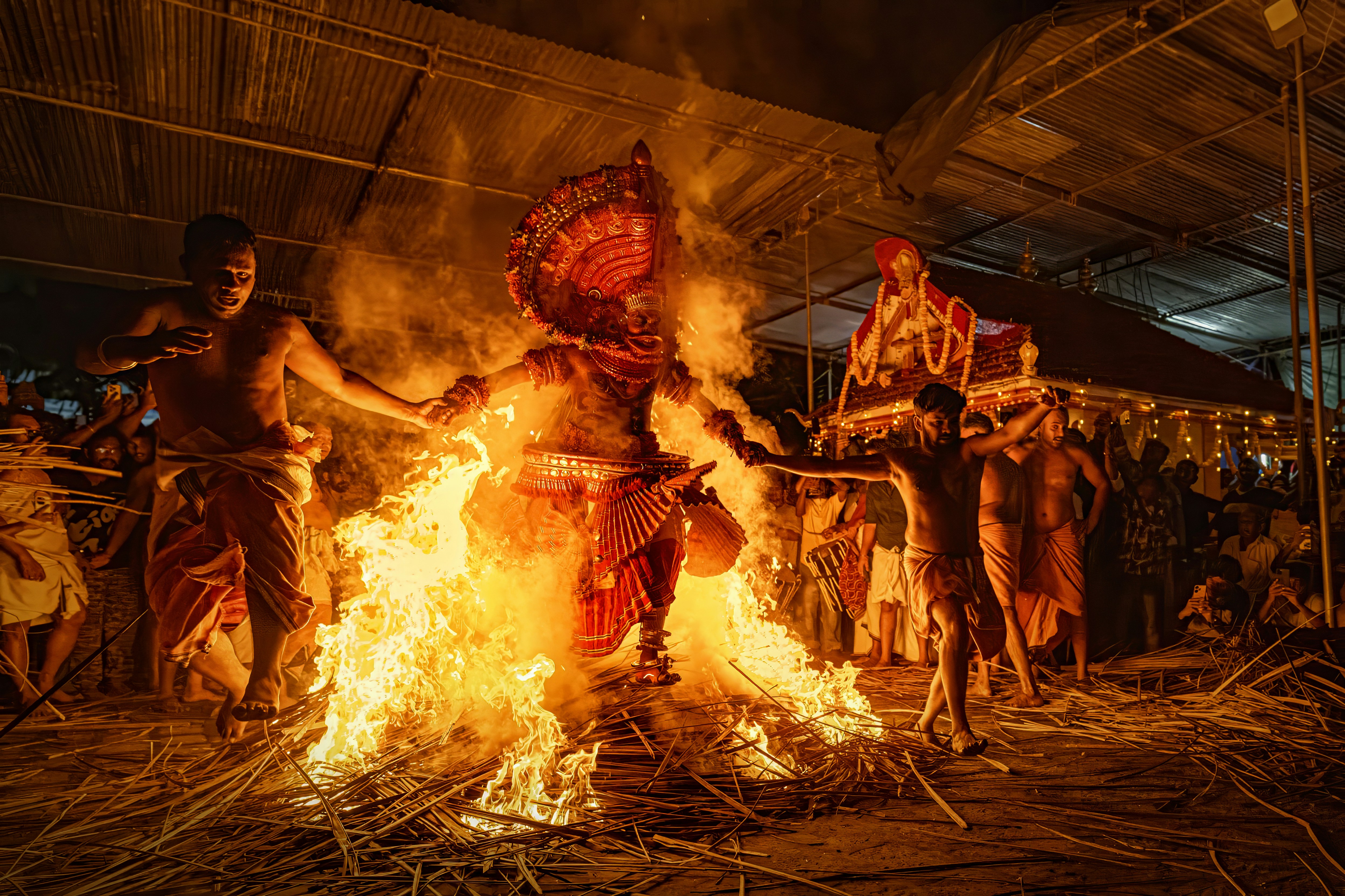 Open-fire ritual with performers around a fierce blaze under a thatched canopy; central dancer in elaborate headgear surrounded by drummers and onlookers, capturing a fiery moment of tradition.