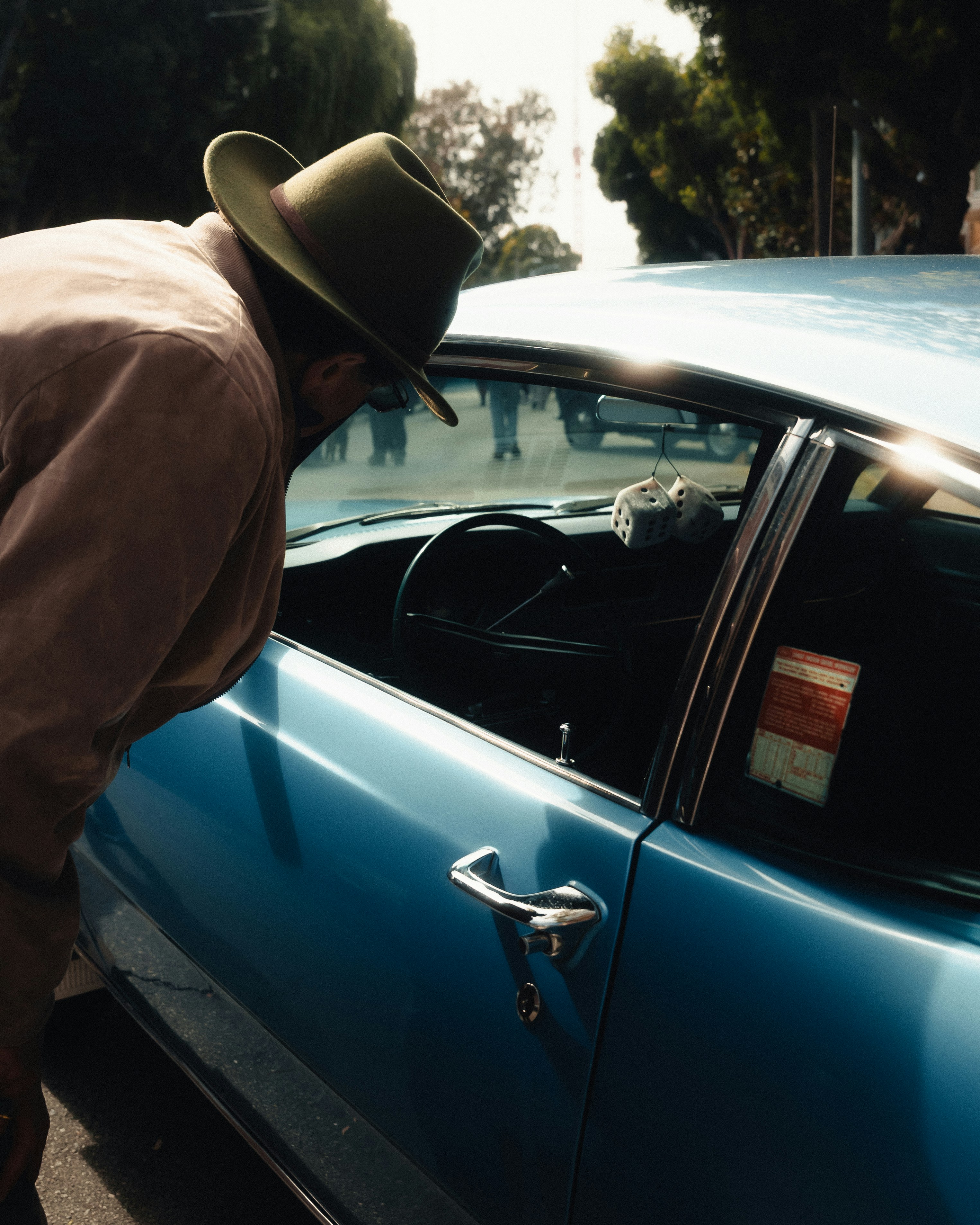 A man wearing a wide-brim hat leans into the open window of a vintage blue car, with a soft, sunlit street scene in the background. It’s a candid automotive moment captured with subtle retro tones.
