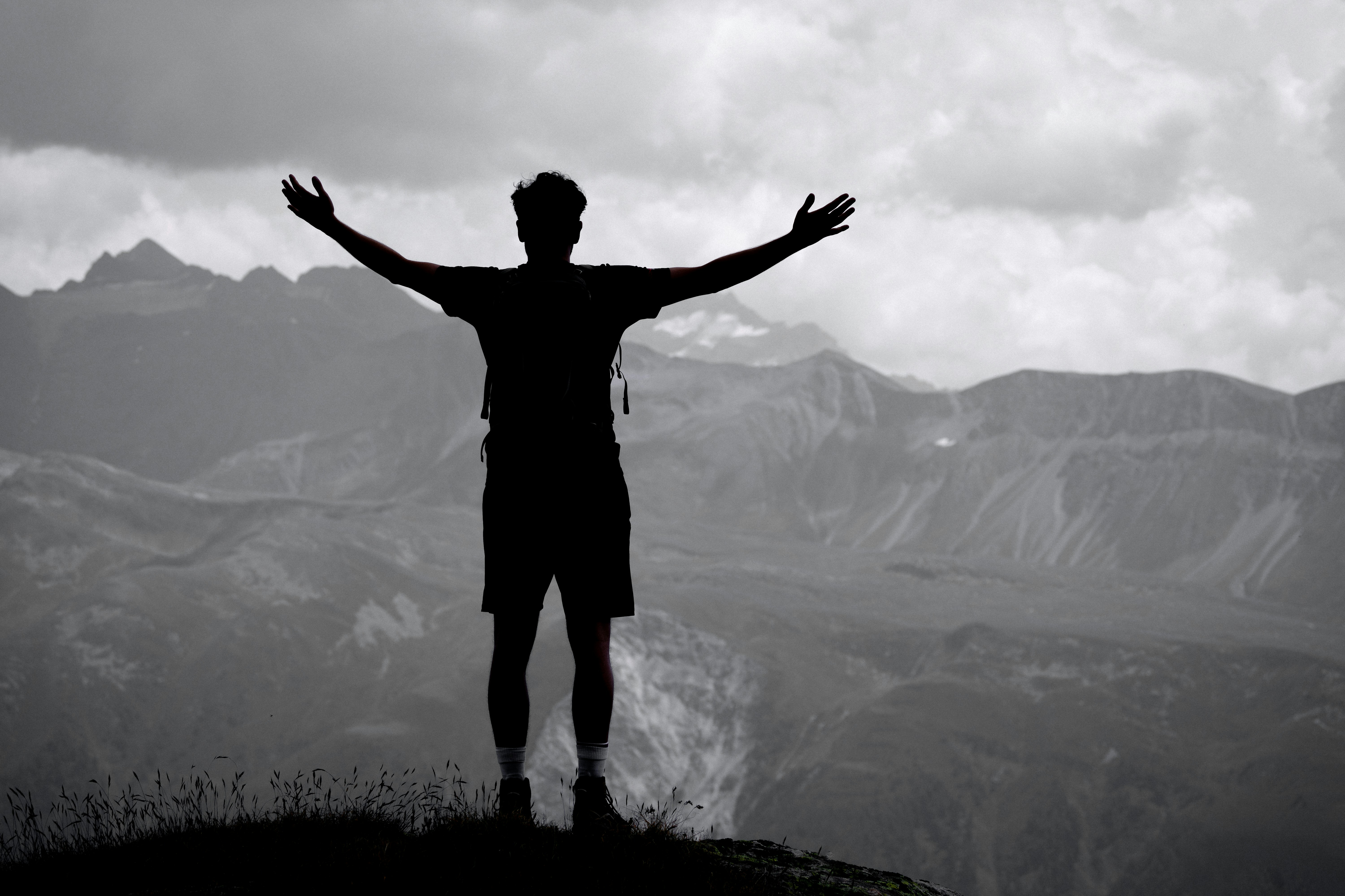 A lone hiker silhouette stands with arms outstretched atop a ridge, against a broad mountainous backdrop. The scene emphasizes scale and solitude, with the figure sharply contrasted in black against a pale landscape.