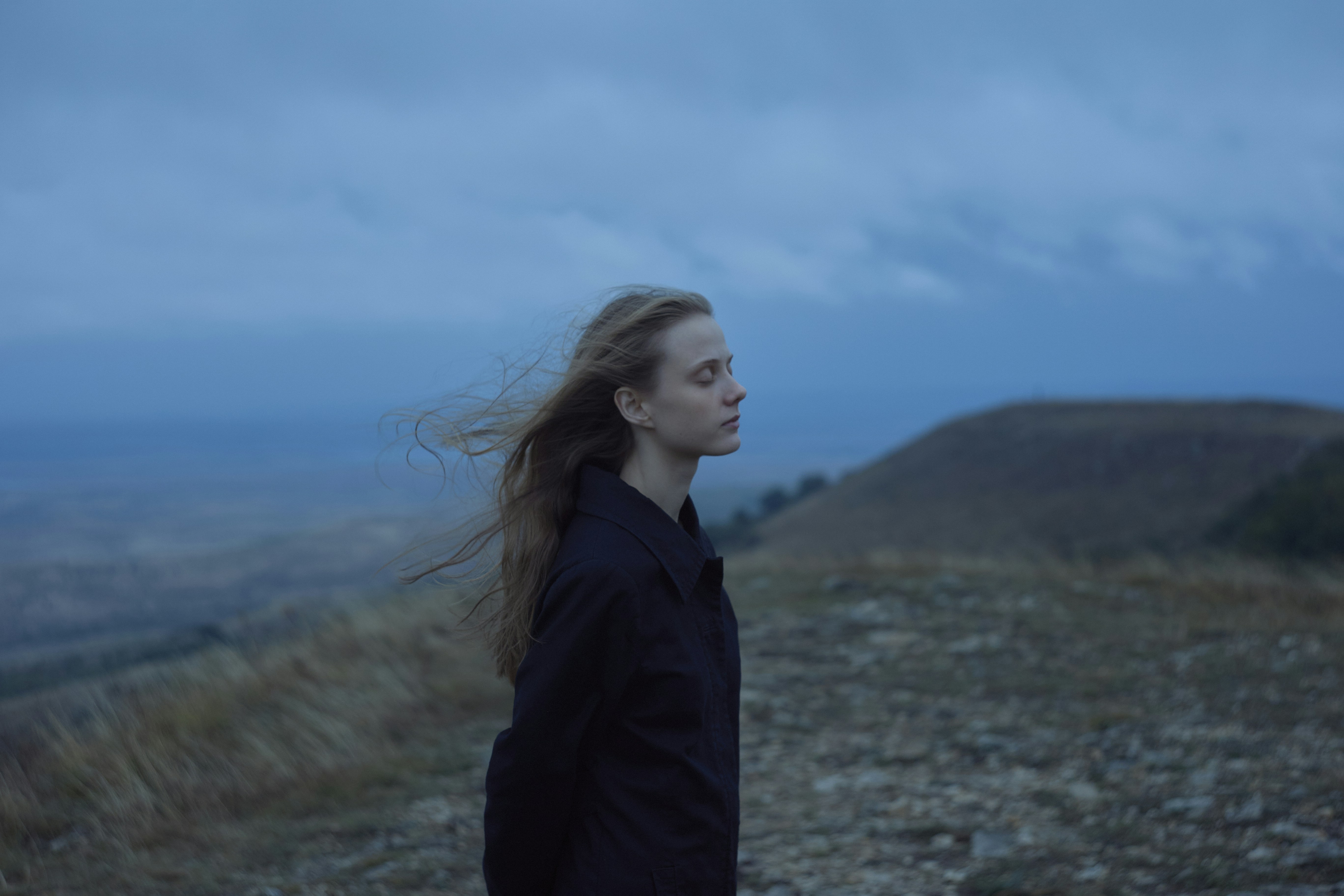 A solitary woman stands at a windswept hilltop, gazing toward a distant horizon; a portrait with a moody blue dusk and a rugged path framing the calm scene.