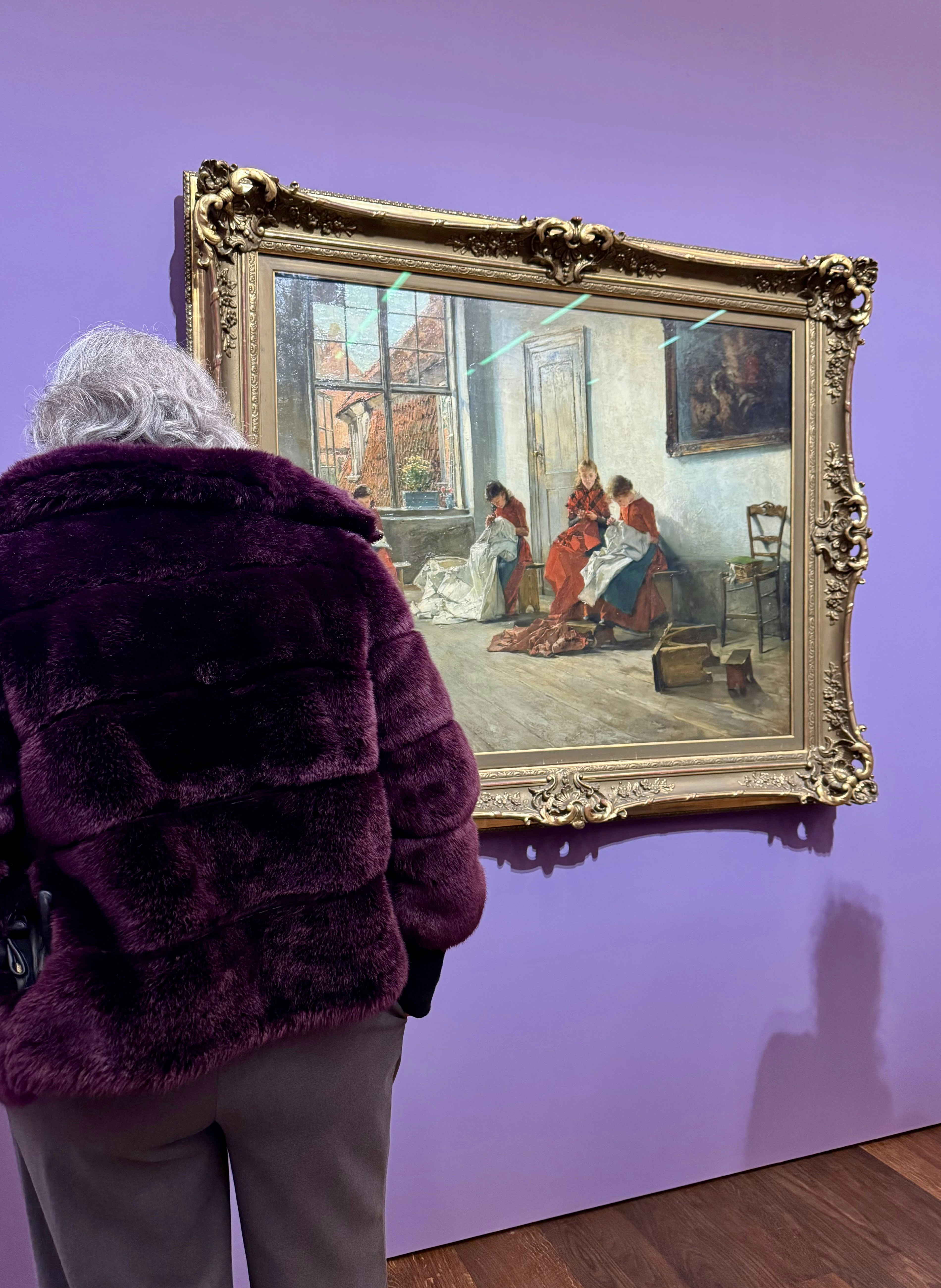 A visitor in a deep purple fur coat stands with their back to the camera, viewing a large framed painting on a lavender wall in an art gallery.